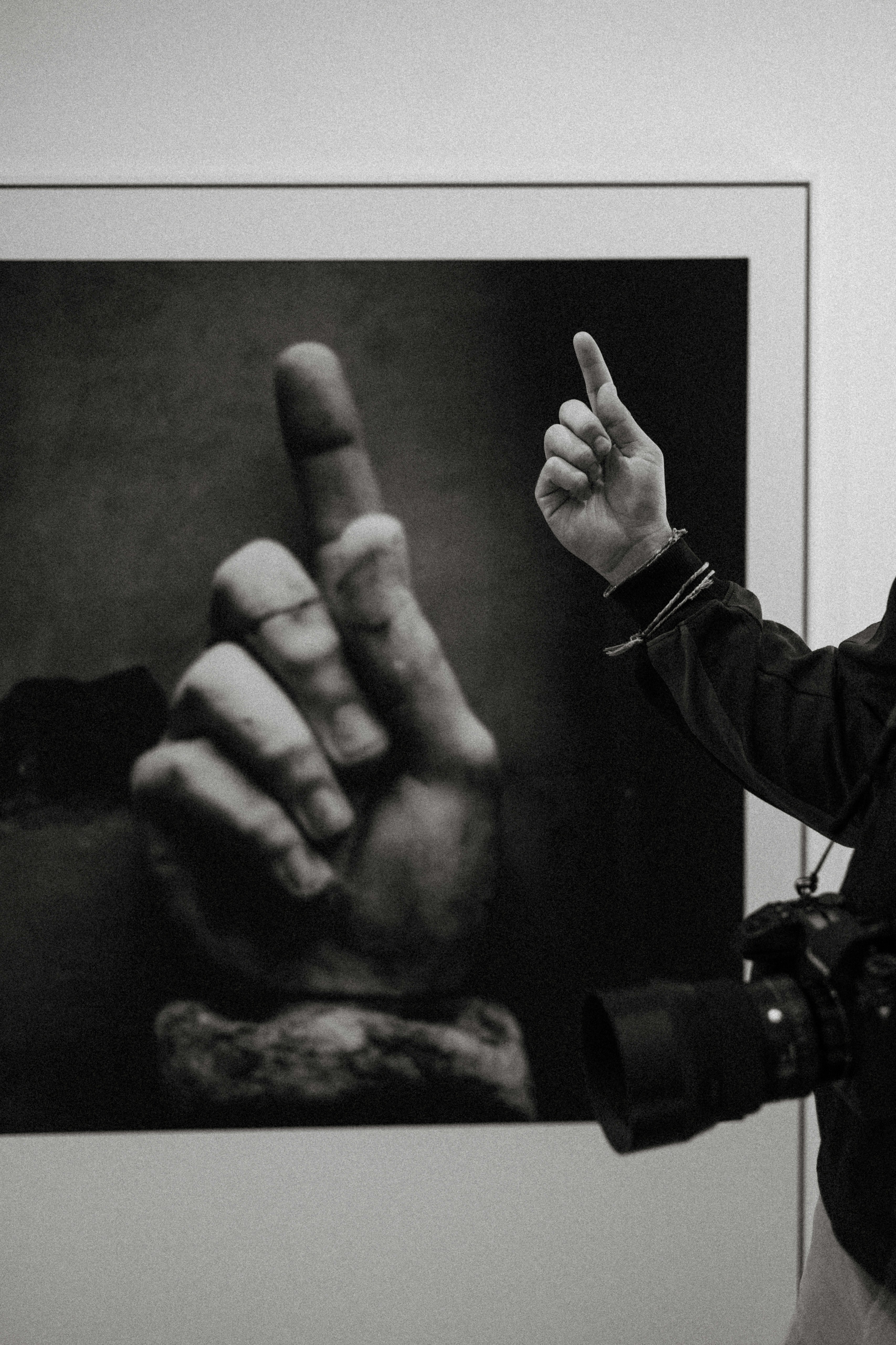 Monochrome scene of a photographer pointing a single finger toward a large framed hand-gesture photograph on the wall, with a camera visible in the foreground. The composition focuses on the interaction between subject and artwork.
