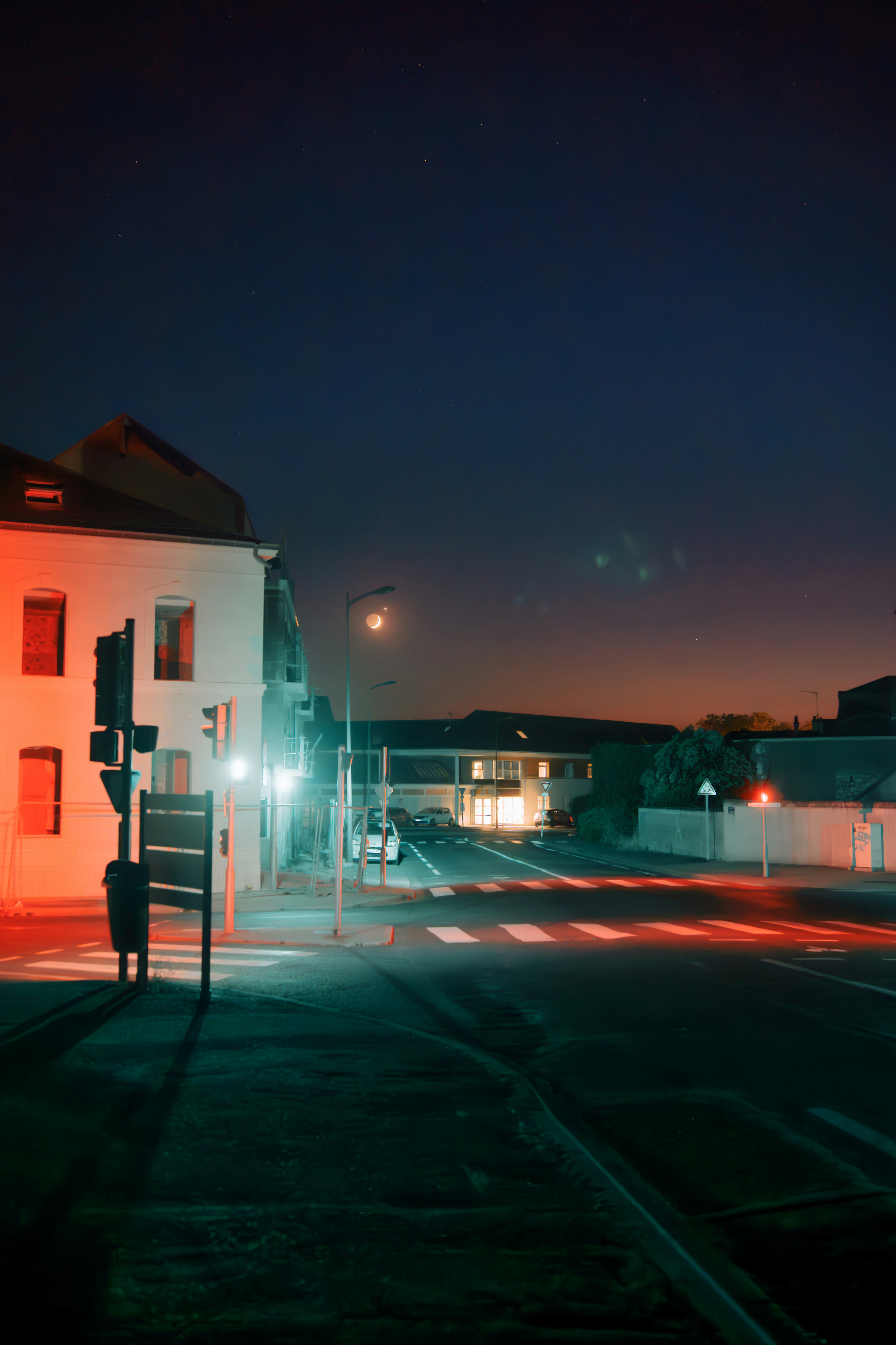Night street scene with a corner building on the left, illuminated by red and teal streetlights; distant cars and a crosswalk recede into a quiet town at dusk.