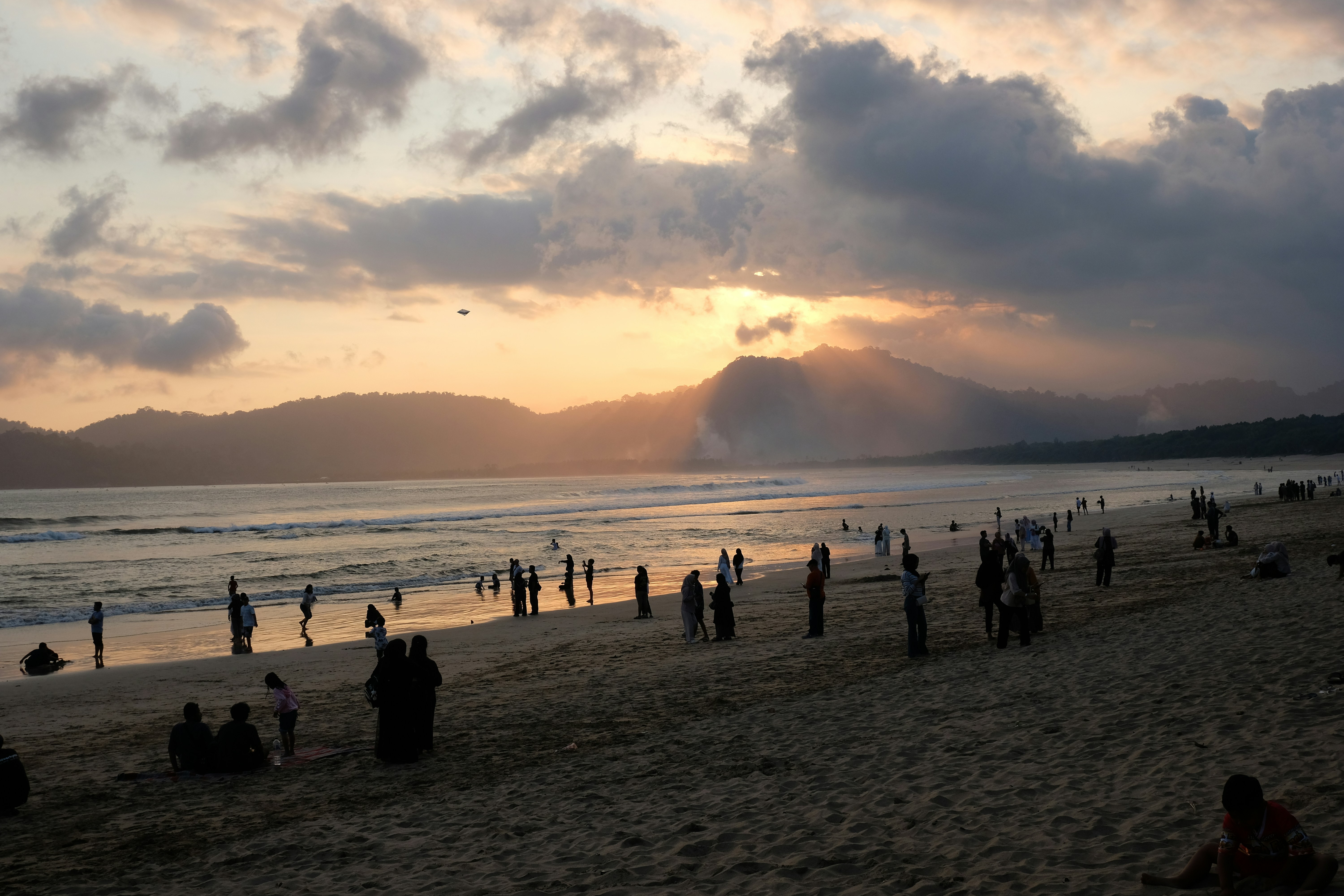 Dusk settles over a crowded beach as silhouettes linger along the shoreline with a glowing horizon and scattered clouds in the sky.