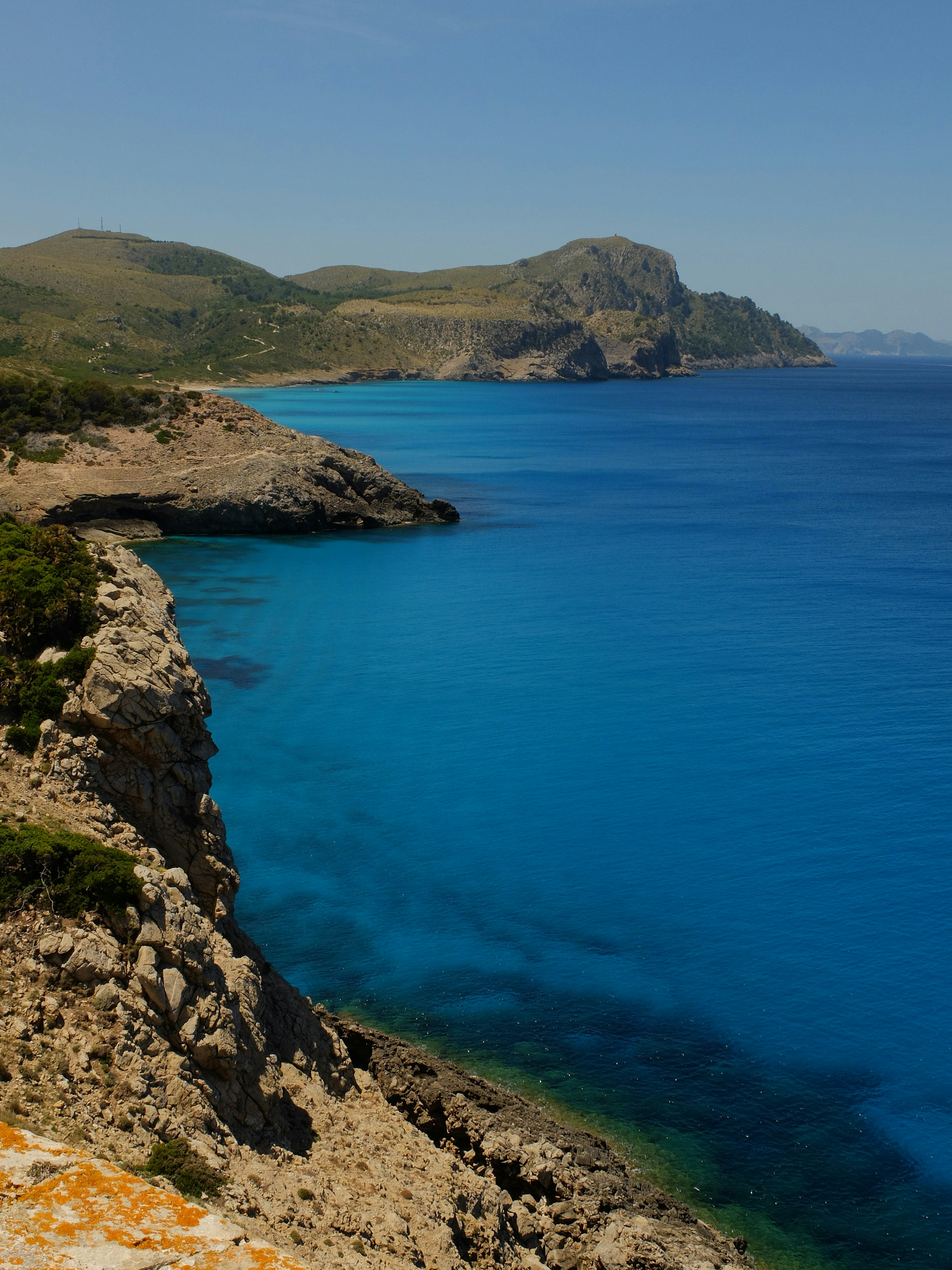 A rugged cliff coastline descends into calm blue water under a bright, cloudless sky; the scene emphasizes the contrast between rocky shore and sea. This photograph appears as a natural landscape capture.