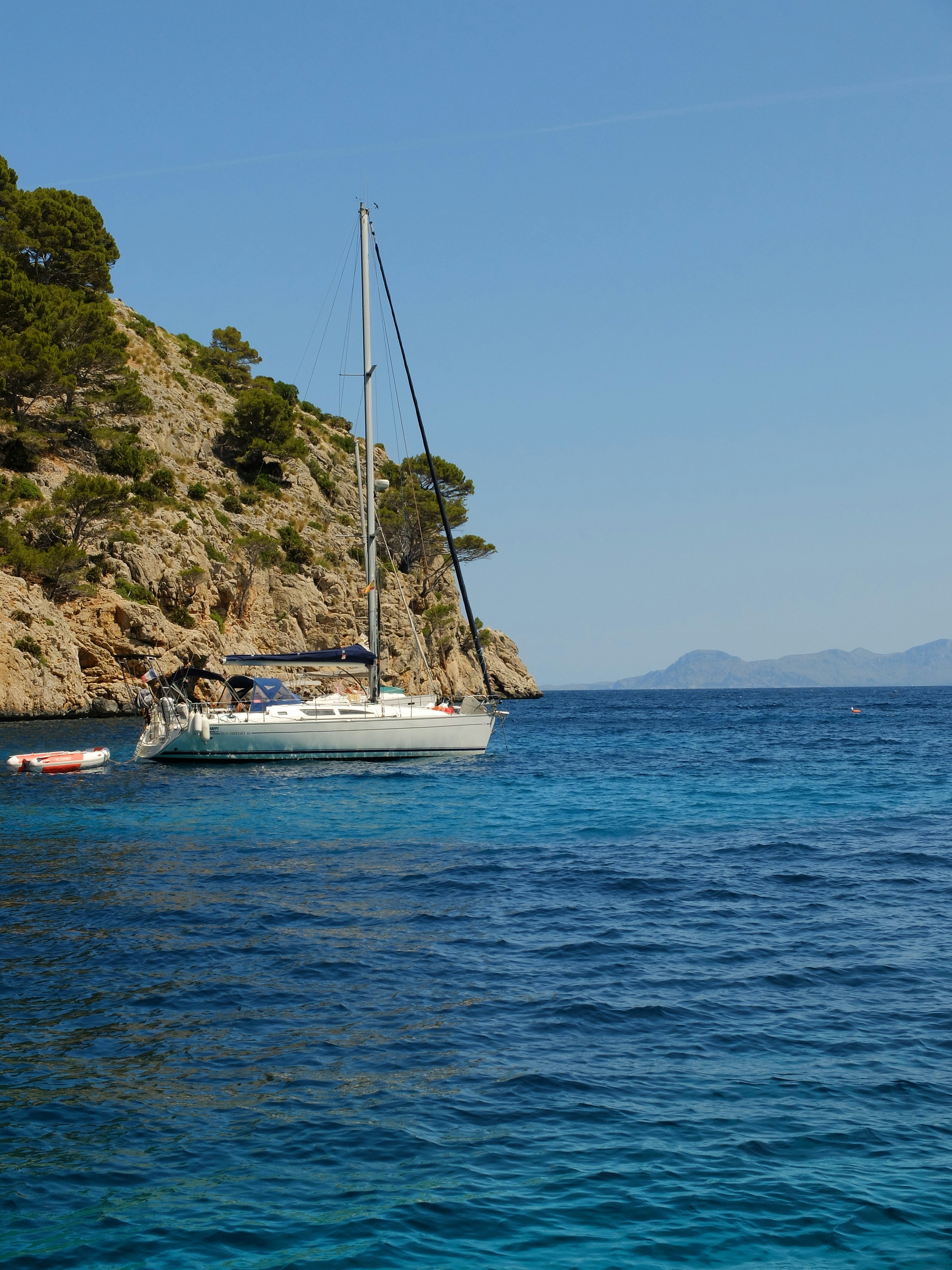 White sailboat rests near a rocky coastline under clear blue sky, with calm turquoise water surrounding the scene.