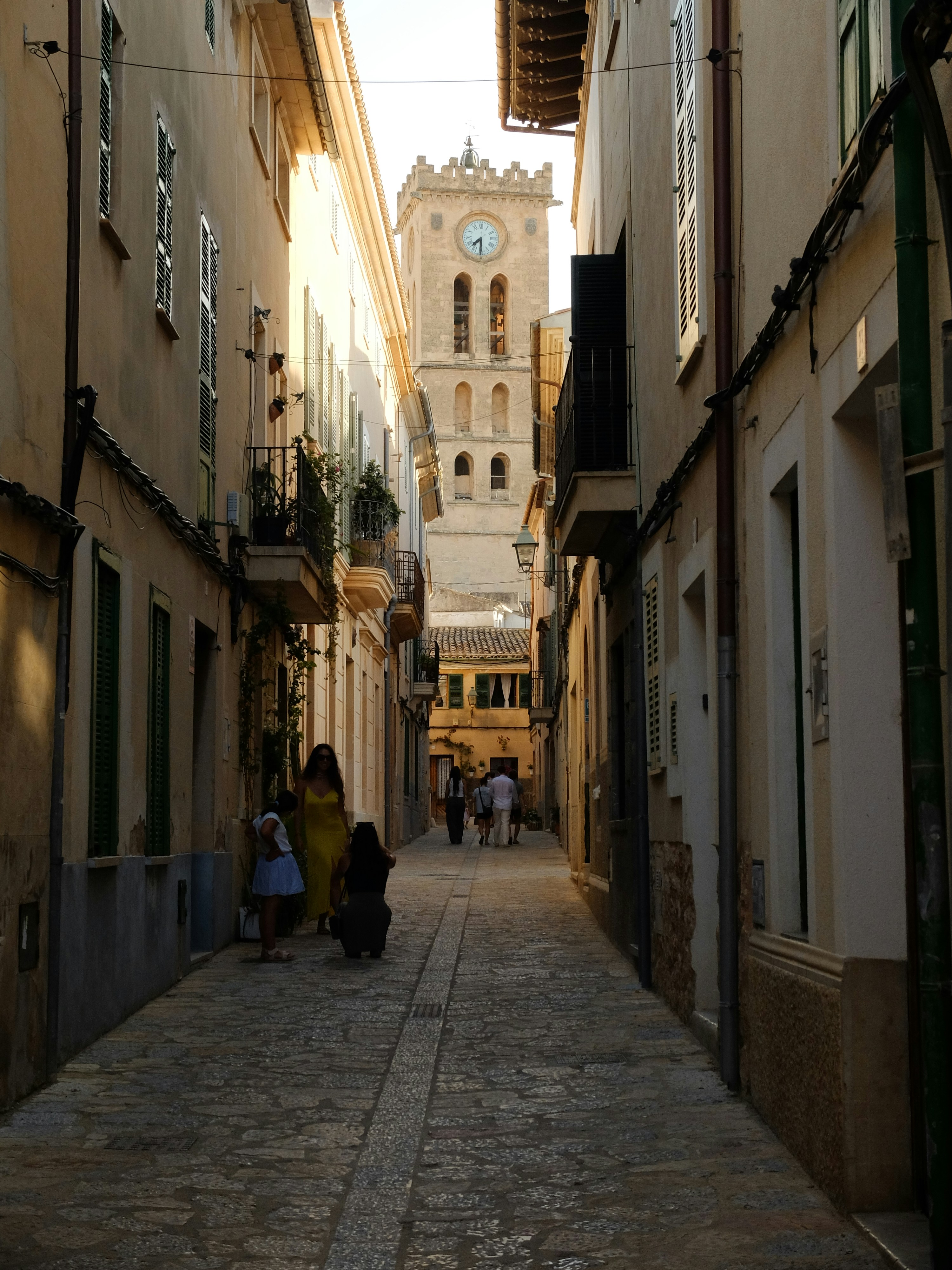 Sunlit medieval alley leads toward a clock-tower facade, with residents strolling and warm tones along rough cobblestones.