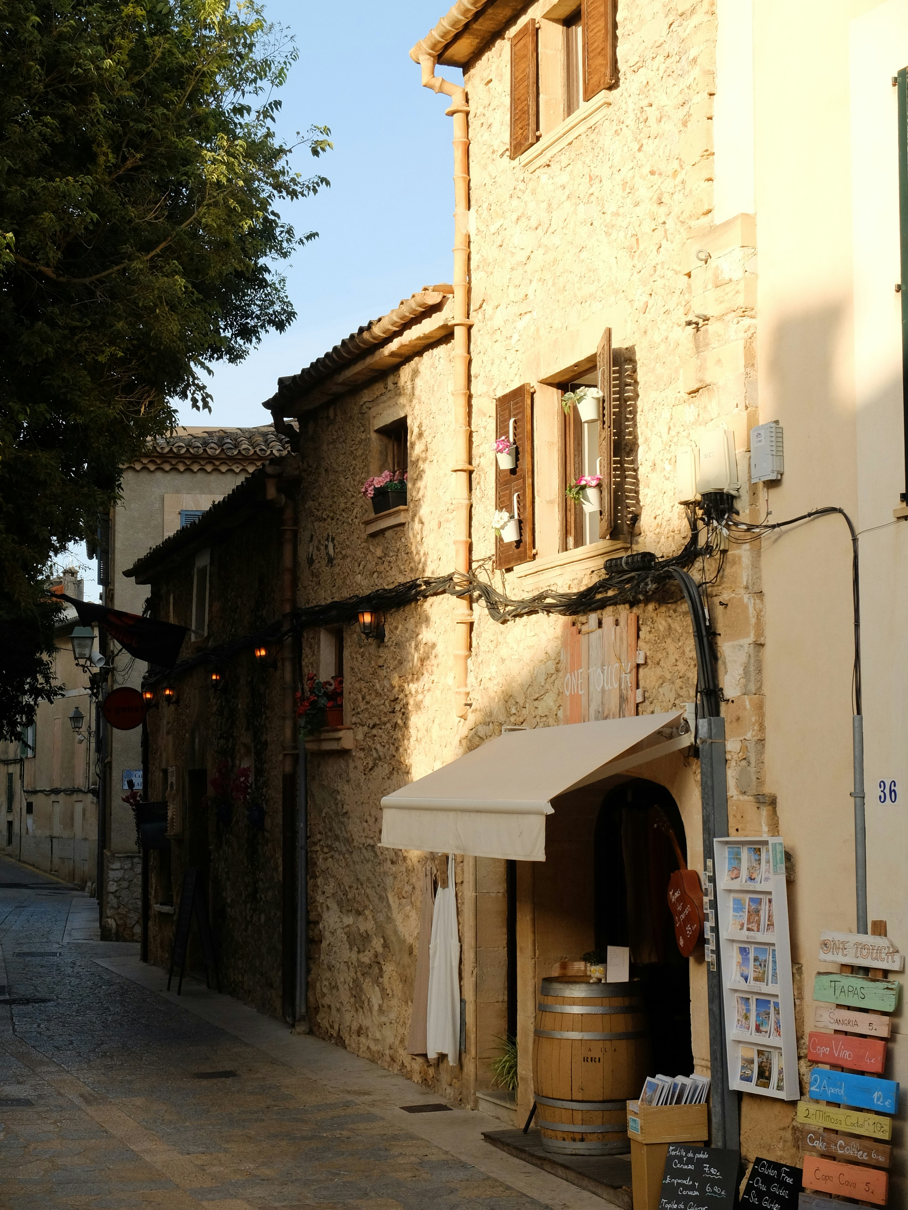Sunlit stone buildings line a narrow street with a small café awning and wooden barrels, evoking a quaint, sun-warmed village scene.