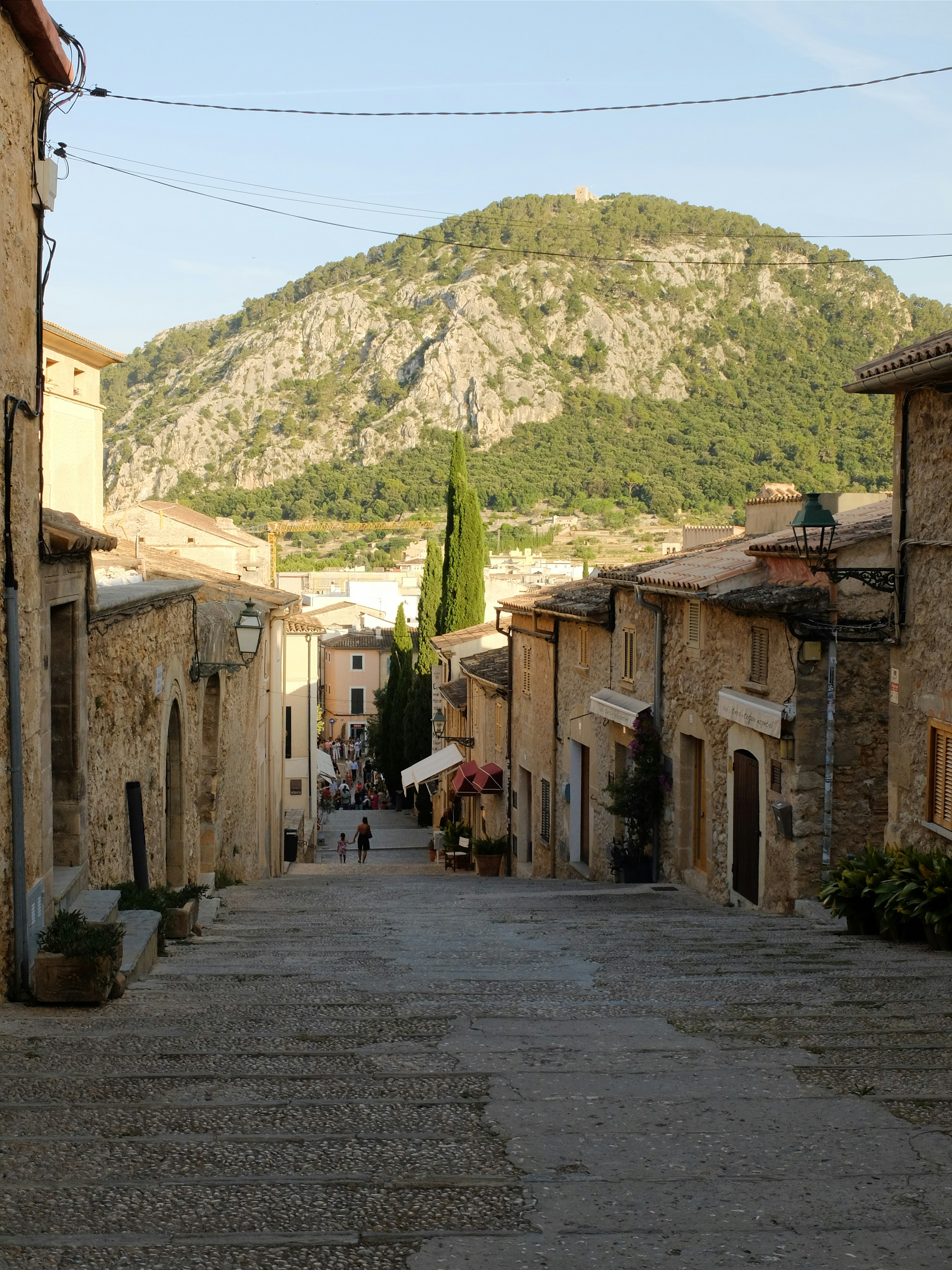 Stone-paved street leads toward a sunlit hillside village with pastel houses and a distant mountain; pedestrians add life to the quiet scene.