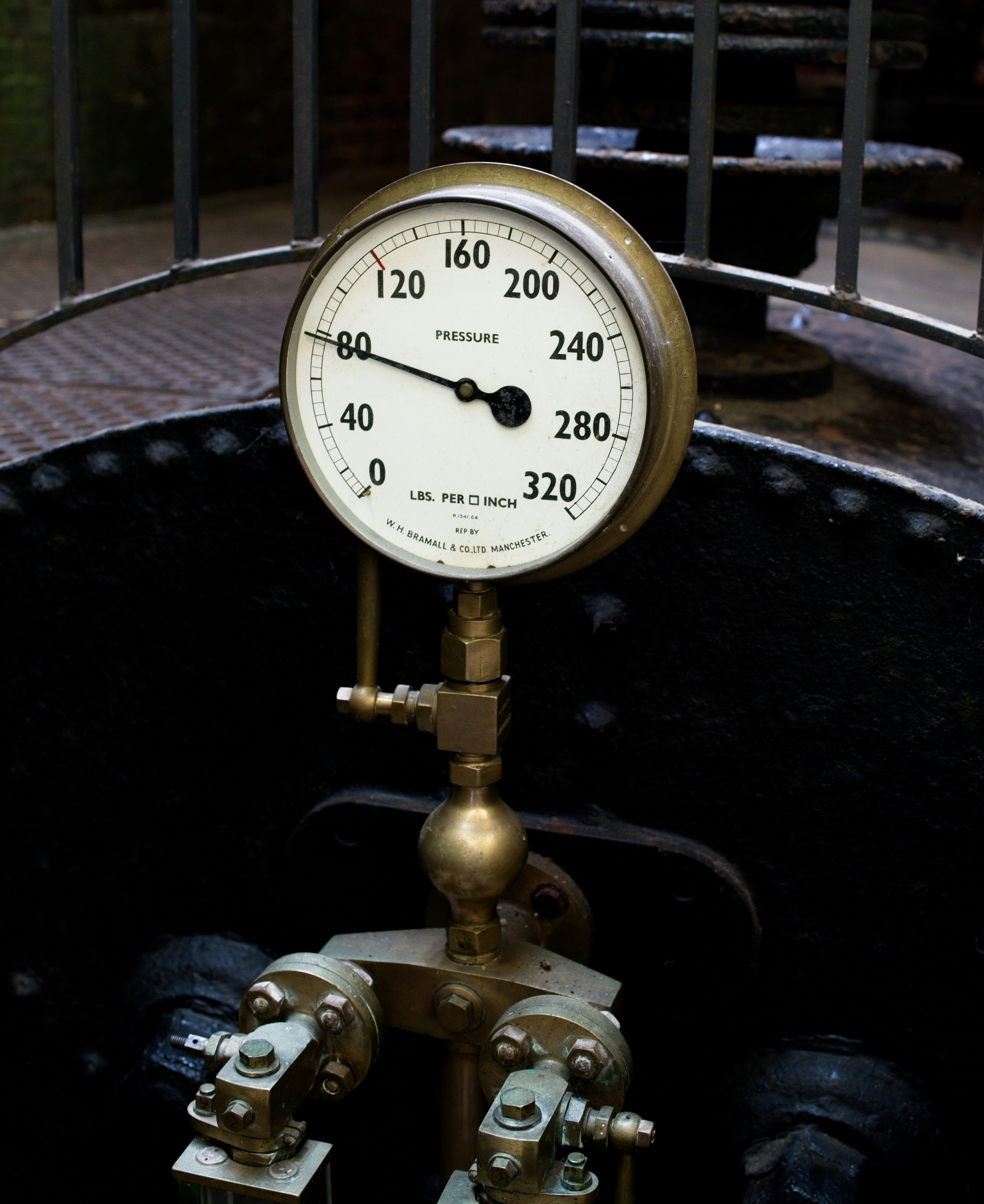 Circular pressure gauge atop brass machinery, framed by dark industrial backdrop and railings. A monochrome, high-contrast instrument with visible scale sits as the focal point in a dim workshop setting.
