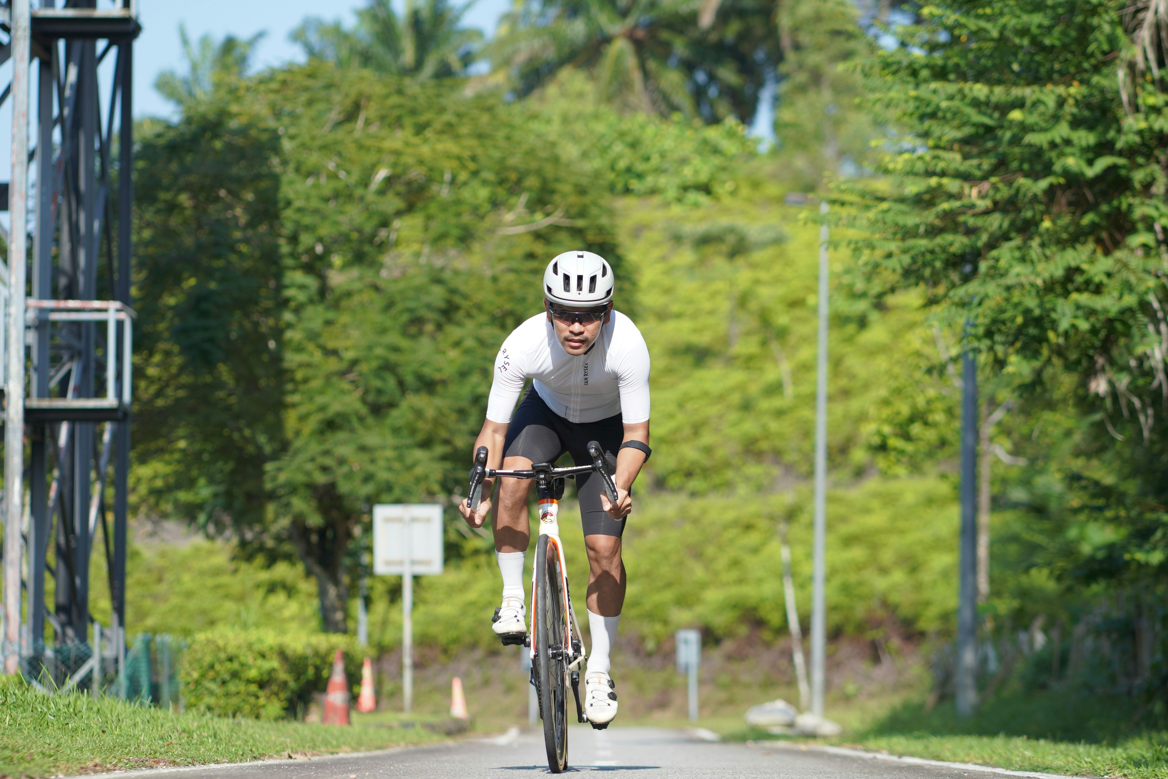 A bicyclist in white gear sprints toward the camera on a tree-lined park road, captured with motion and a sense of speed. This is a photograph.