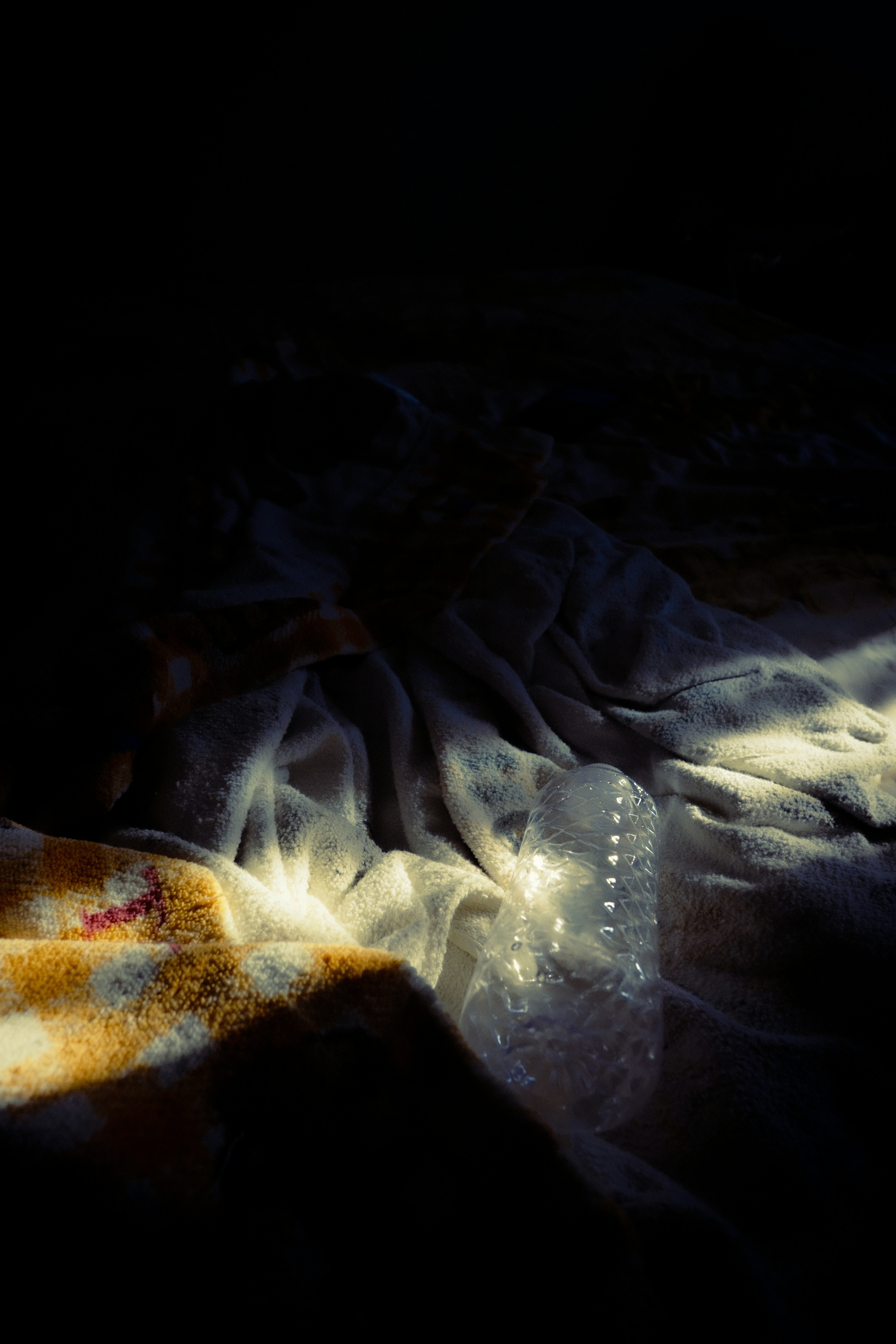 Shadowed Comfort: A Crumpled Blanket Embraces a Hidden Bottle in Dim Light