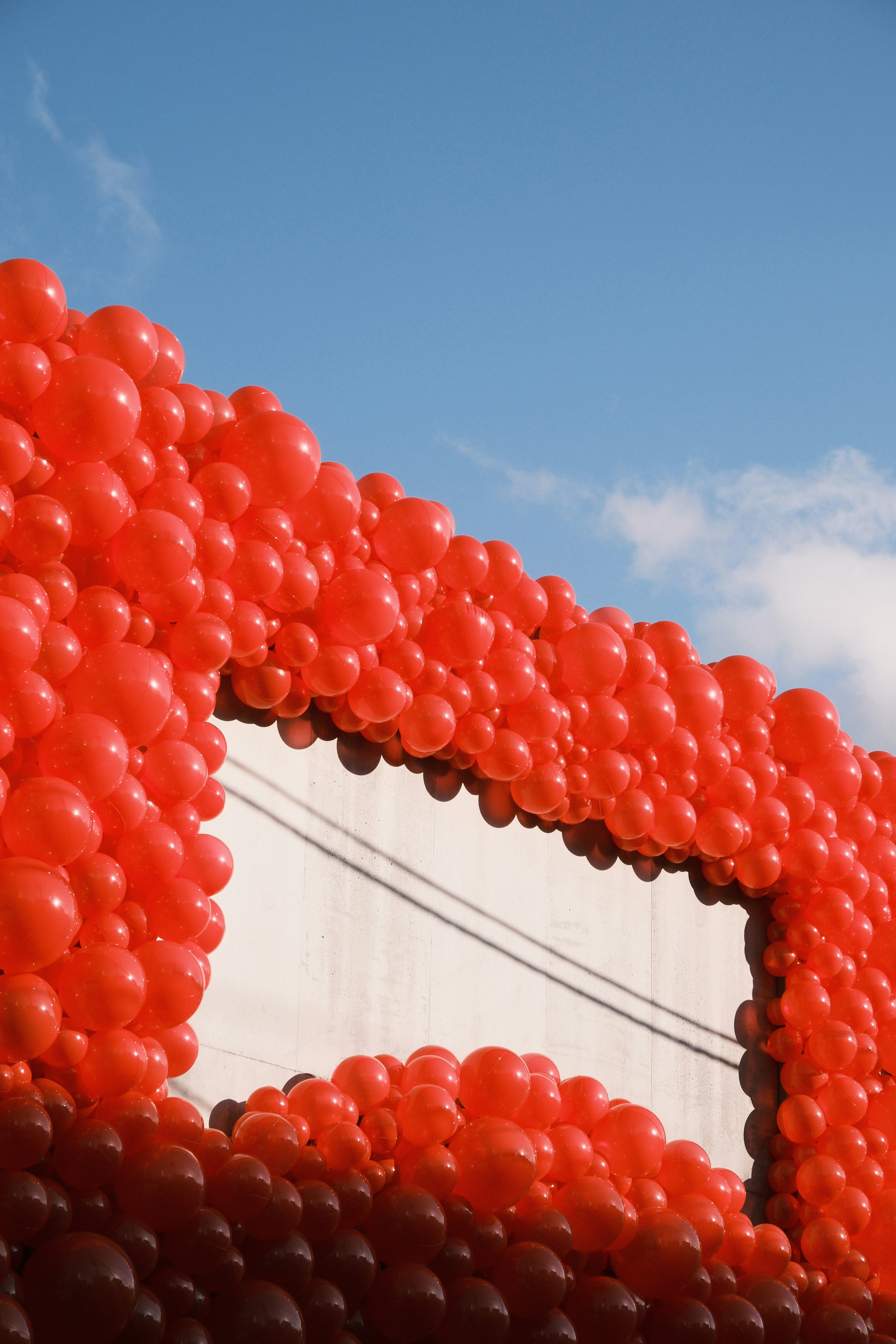 A large archway of bright red balloons frames a white wall under a blue sky, creating a bold, festive installation.