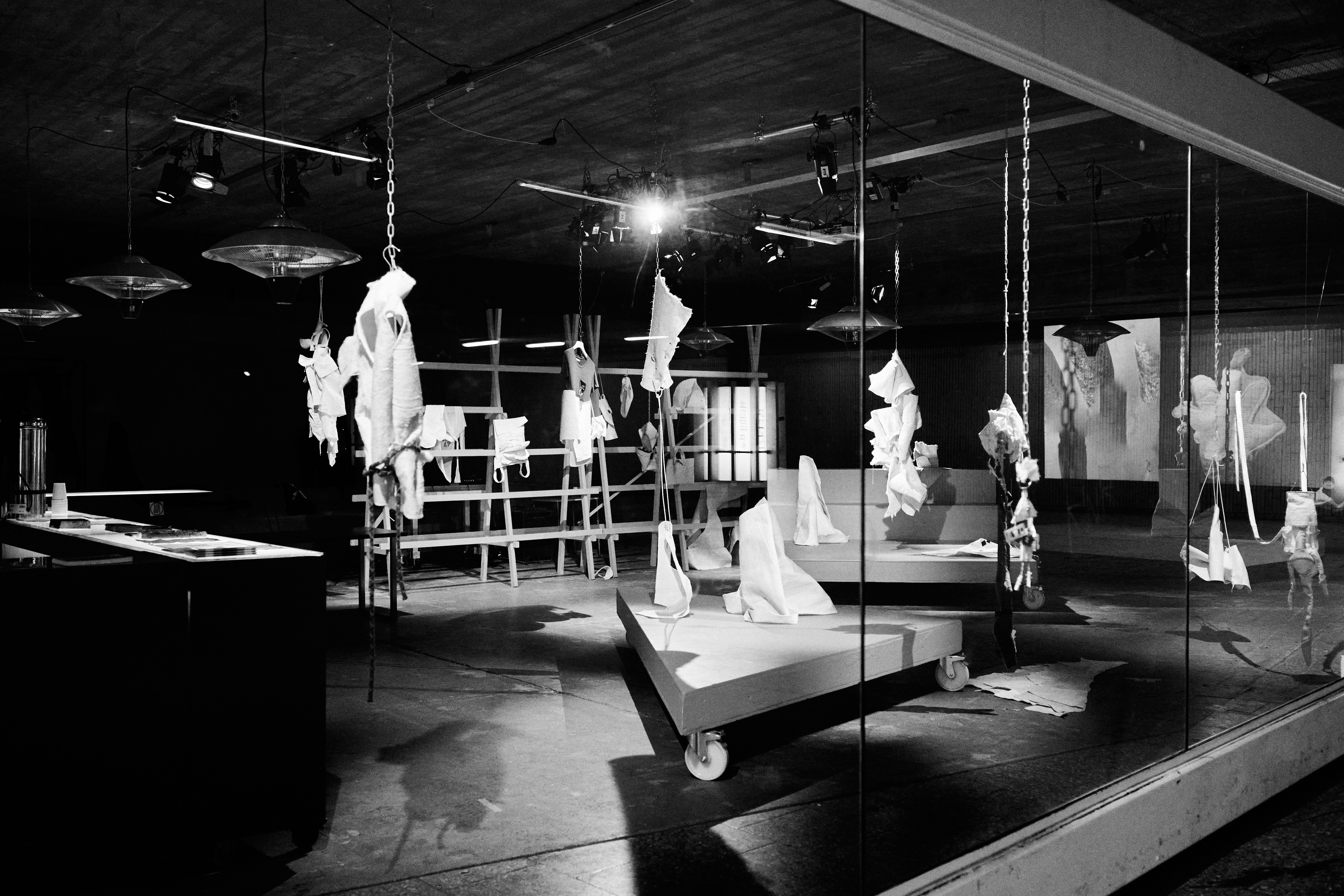 Industrial studio scene with hanging fabric shapes and wheeled platforms, rendered in stark grayscale to emphasize sculpture-like props and lighting.