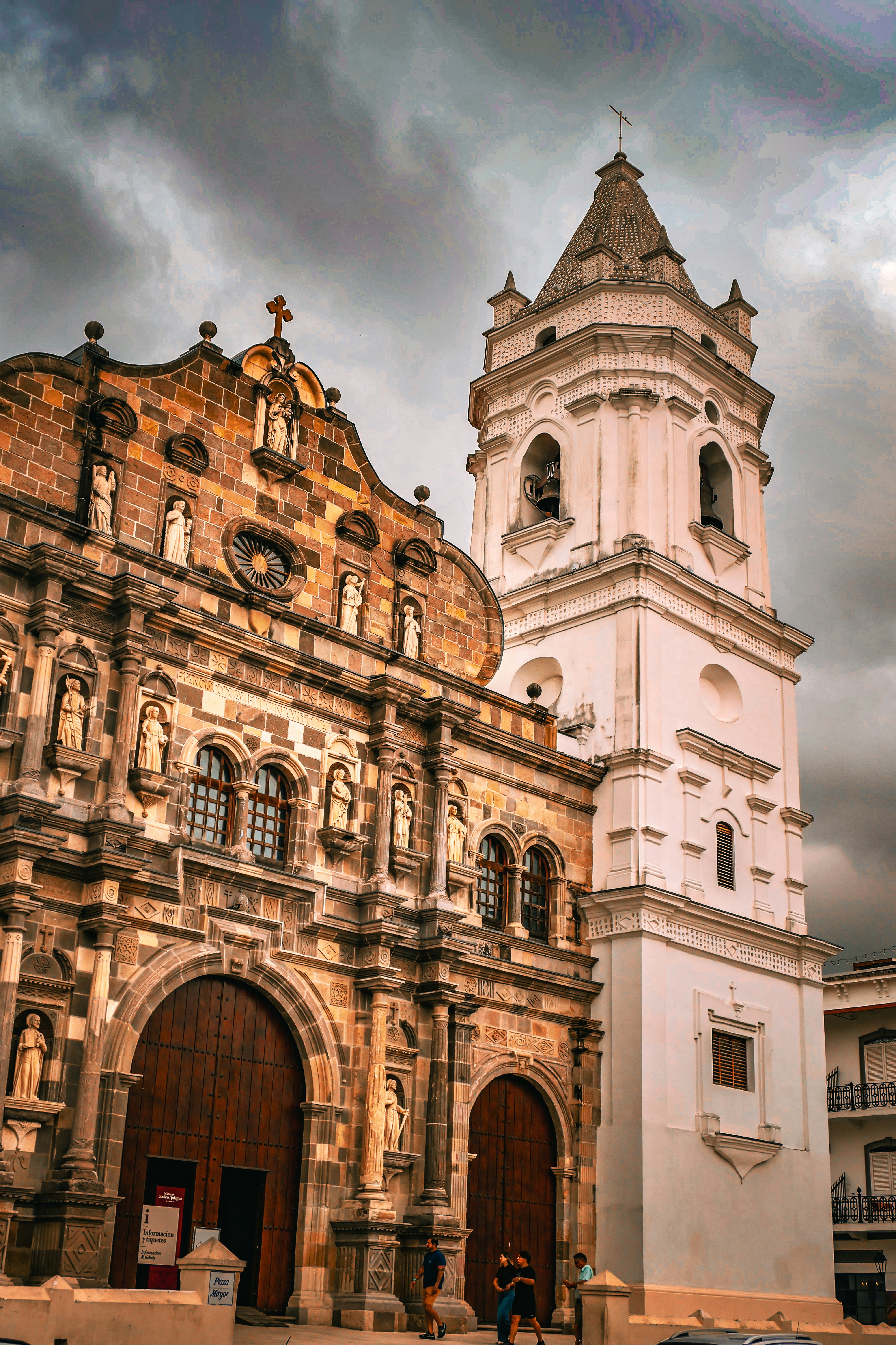 A grand, stone church façade with ornate statues and a white bell tower stands under a moody sky, capturing architectural detail and color. This is a photograph of an exterior building.