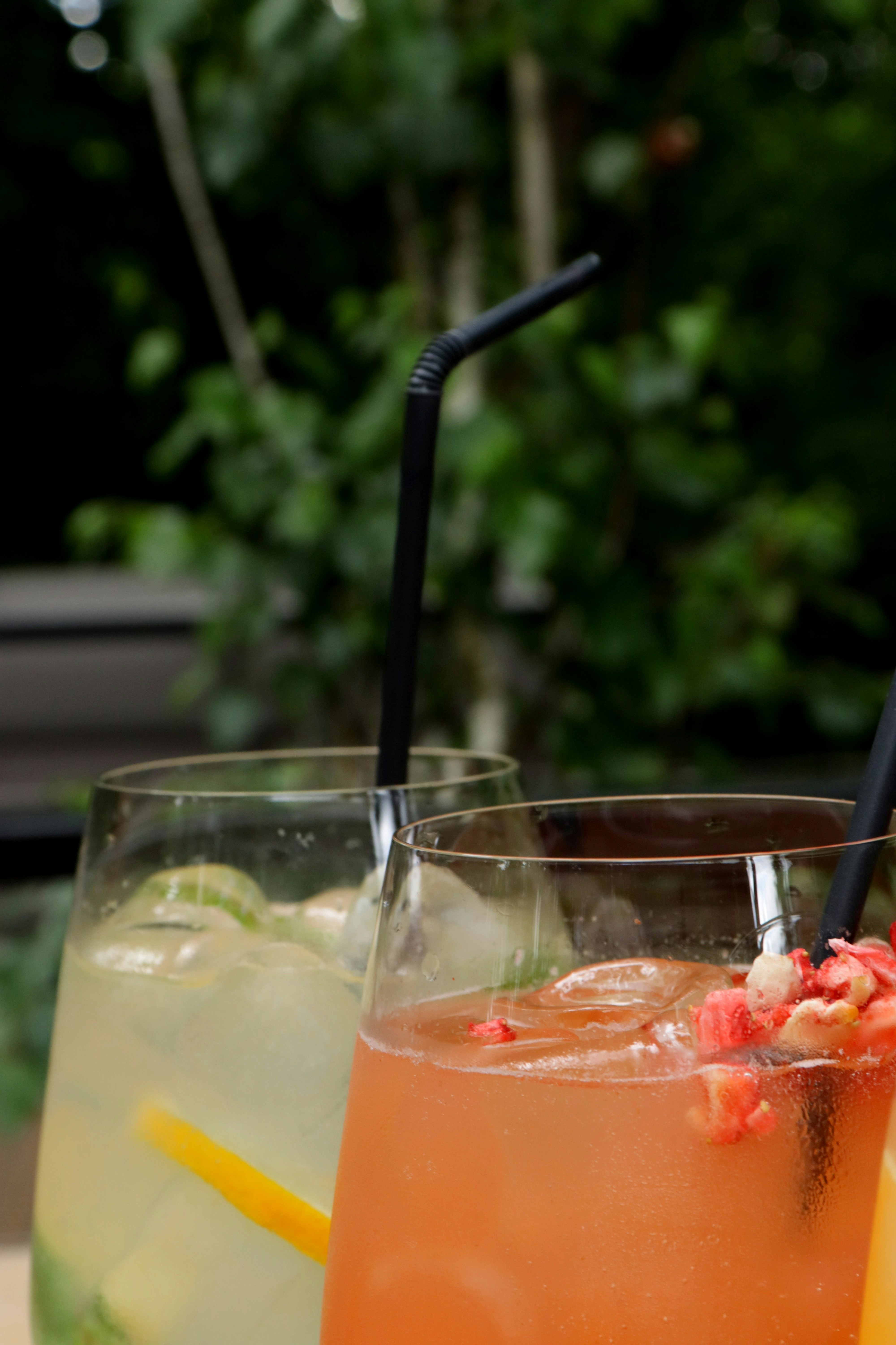 Two cocktail glasses with ice and colorful drinks sit close together on a table, each with a dark straw; a lush green background softly blurs behind them.