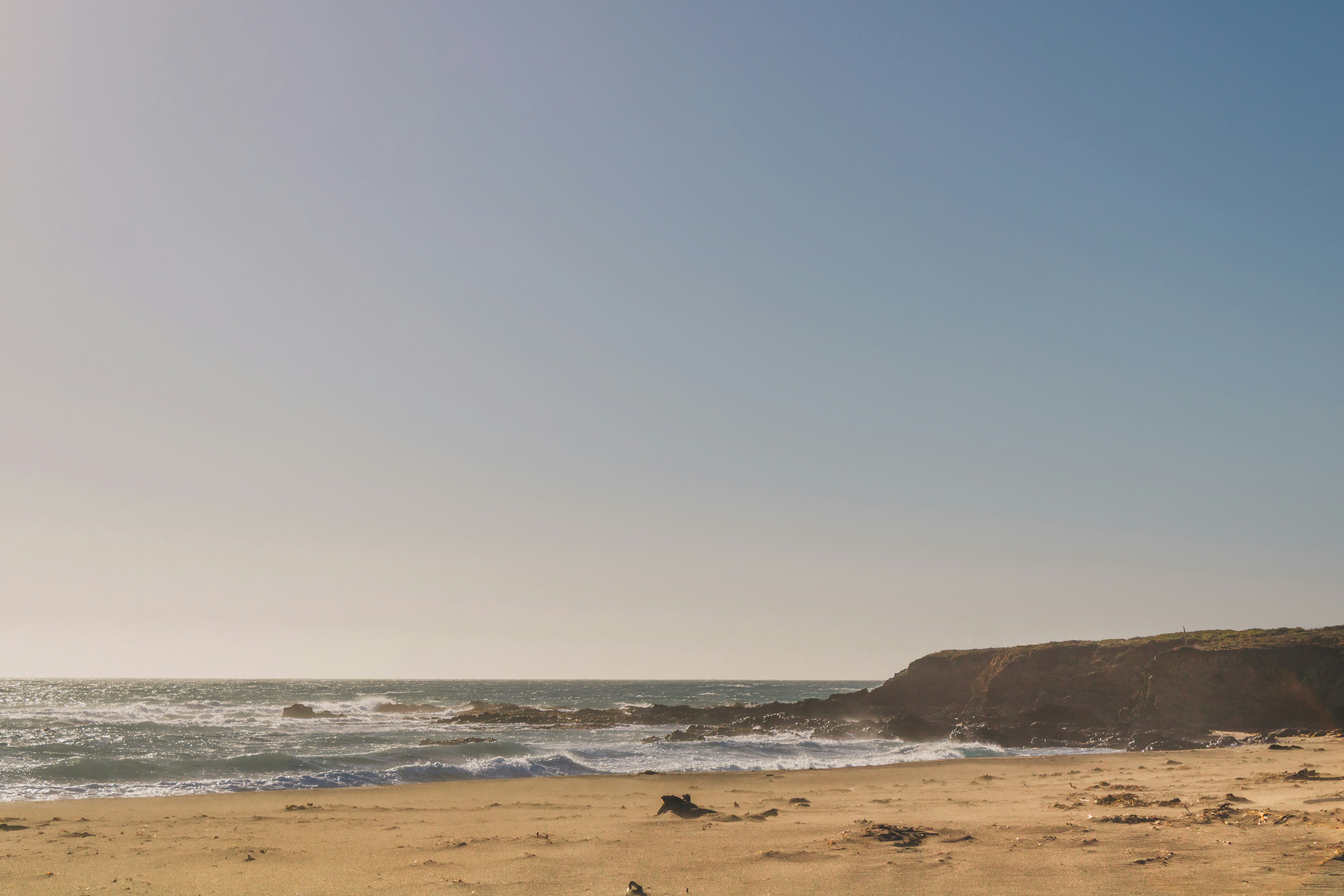 Sunlit coastal scene with a calm sea meeting a sandy shore under a pale, expansive sky; a distant cliff line anchors the horizon.