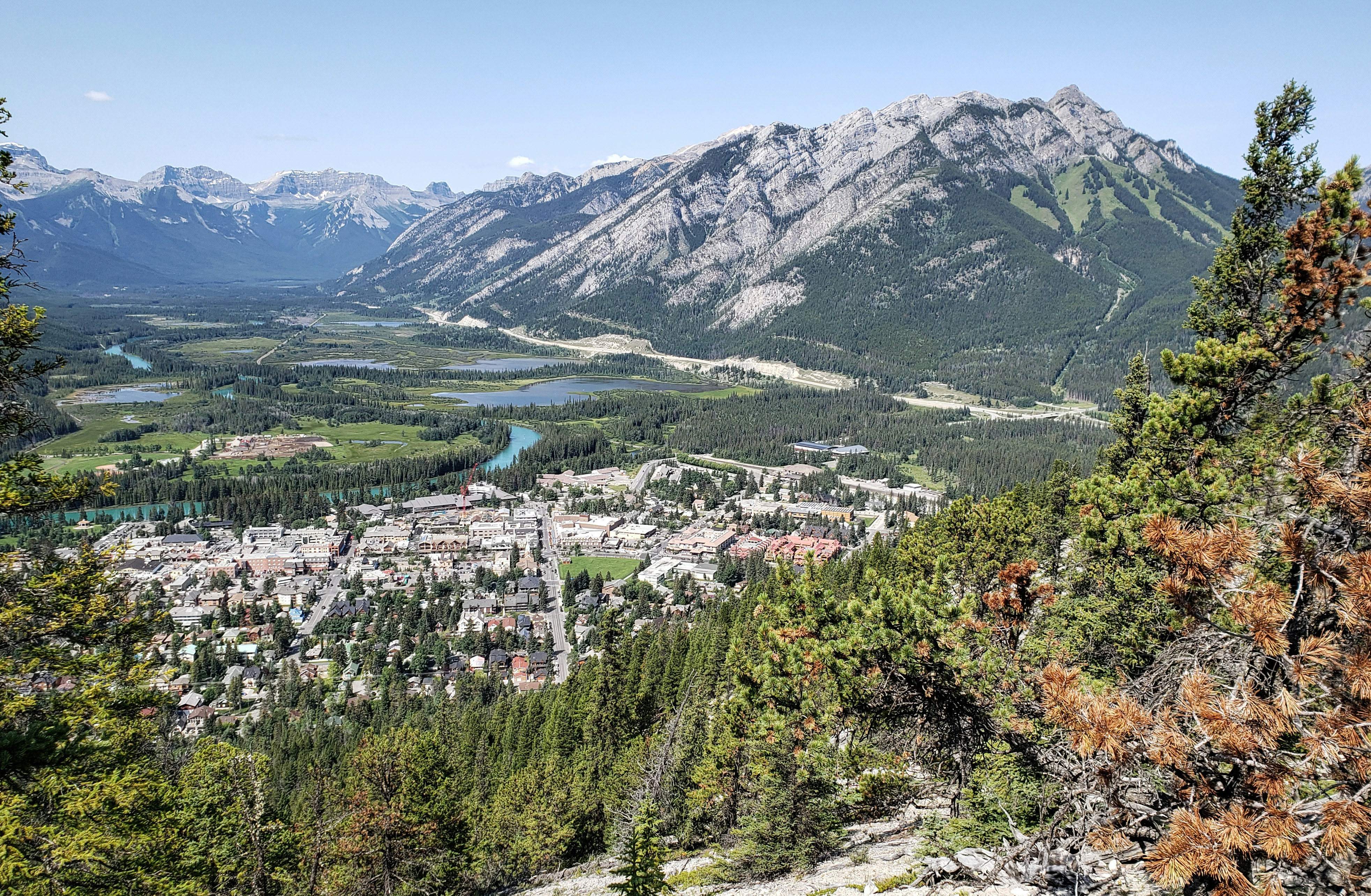 Wide valley with a river loop and a cluster of buildings set beneath rugged mountains; foreground trees frame the town and winding waterway.