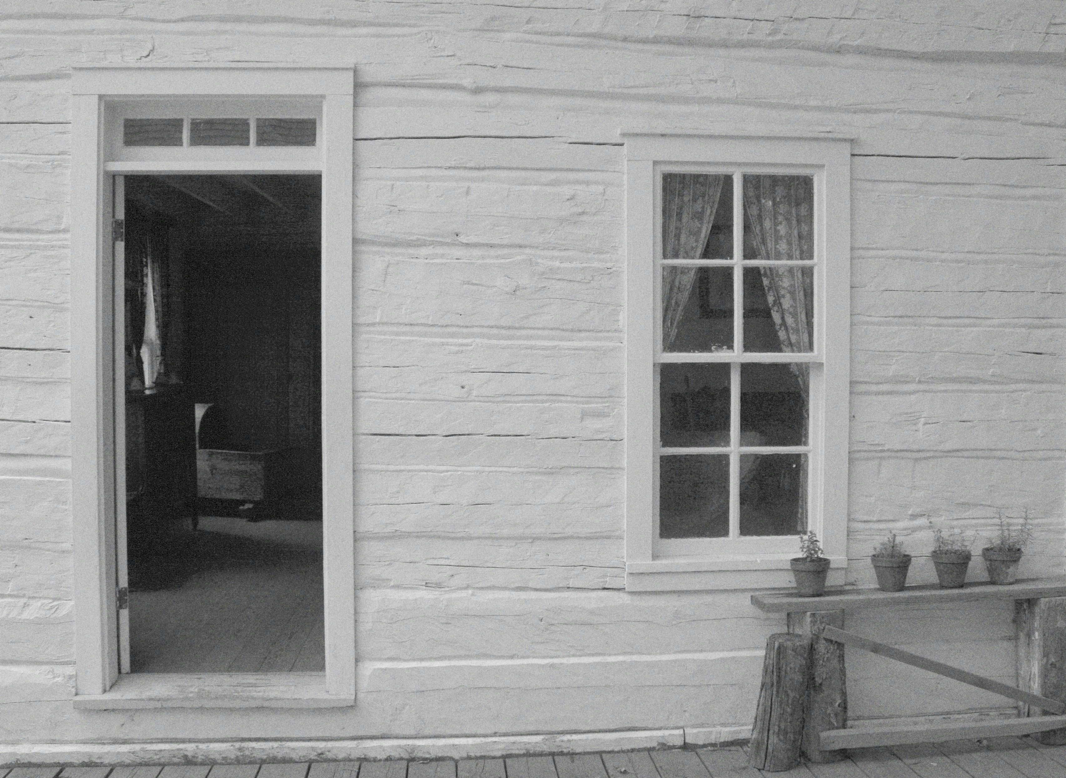A grayscale exterior scene features a weathered clapboard wall with an open doorway on the left and a window with curtains on the right, plus a small row of potted plants on a wooden bench. An understated, rustic still-life of a quiet entrance.