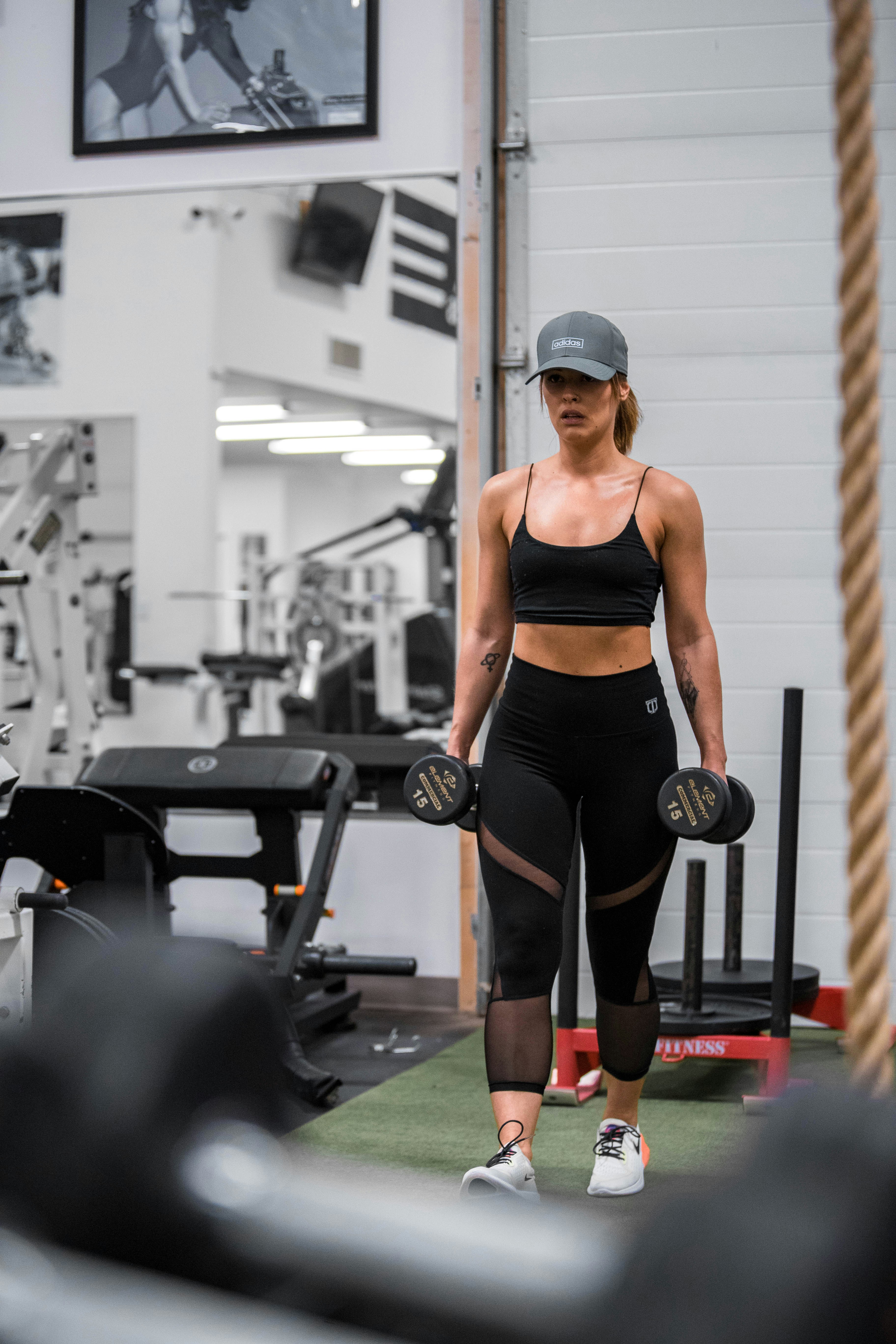 Athlete in black workout gear stands with dumbbells in a gym, preparing for a lift as equipment and mirrors form the backdrop. This is a candid gym scene.