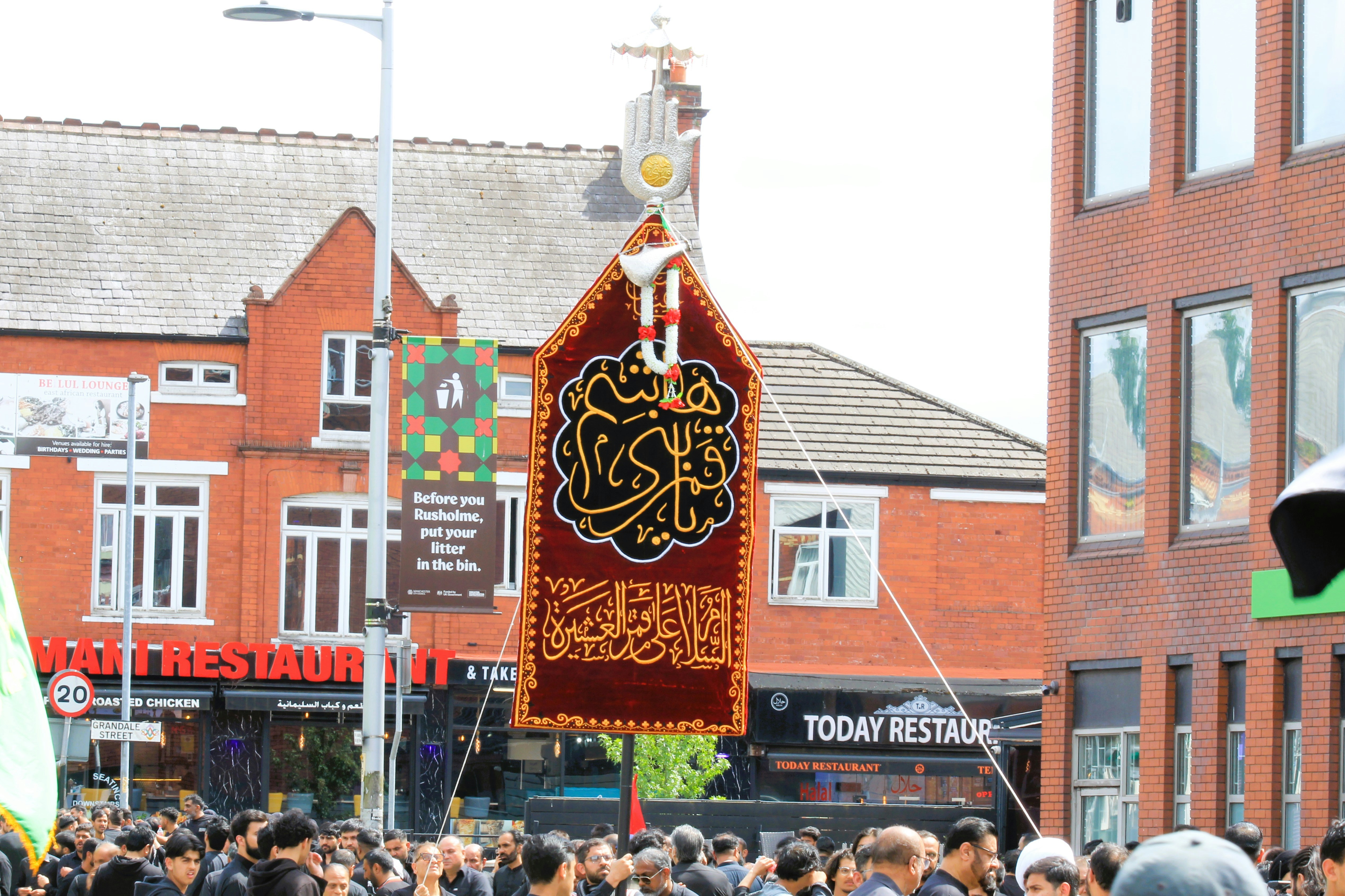 Crowd gathered for a street procession with a prominent ornate banner held aloft amidst urban storefronts.