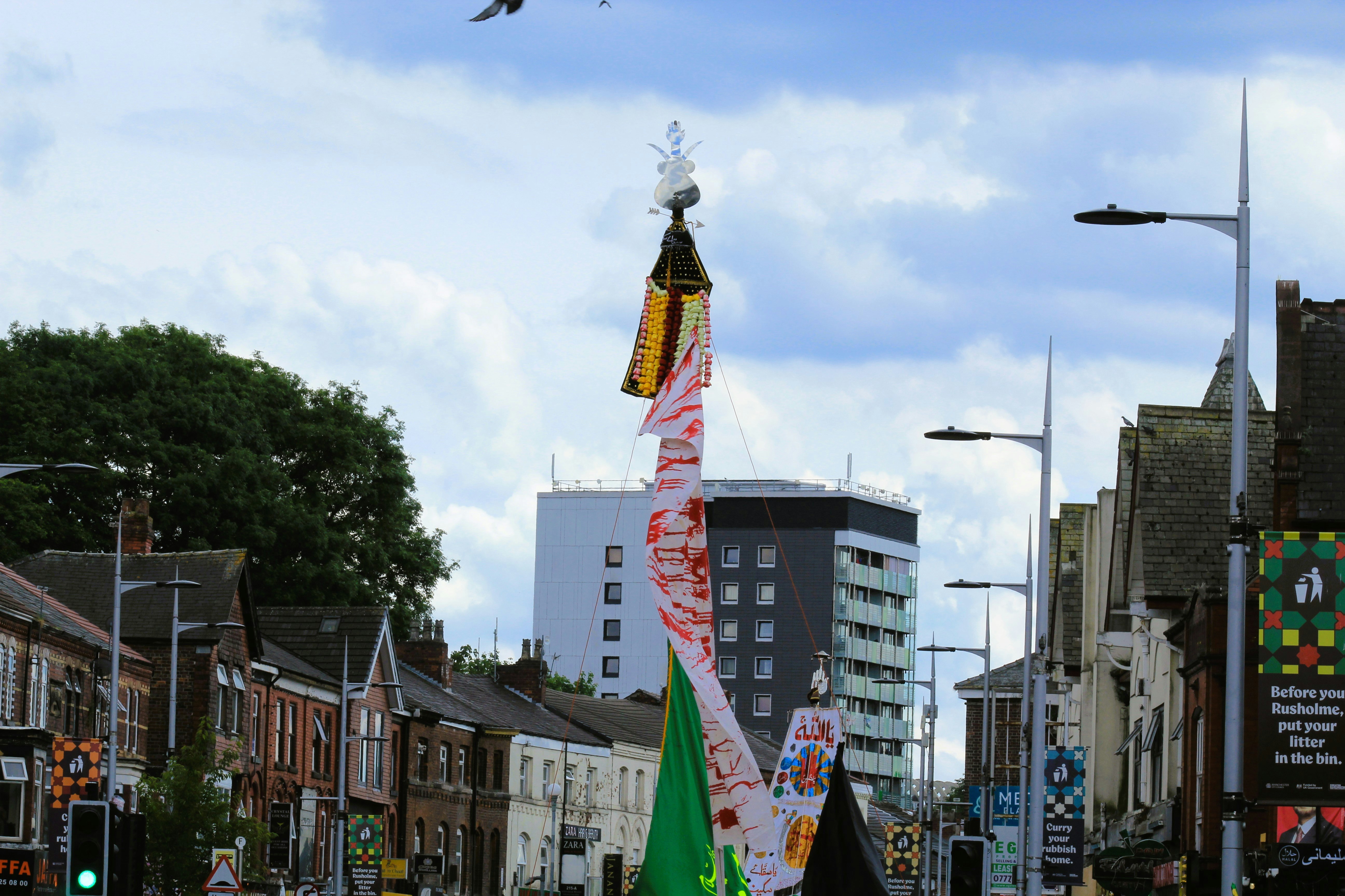 Vibrant festival banners rise along a busy town street, with a tall decorative pole center stage against a backdrop of low-rise buildings and a modern high-rise.