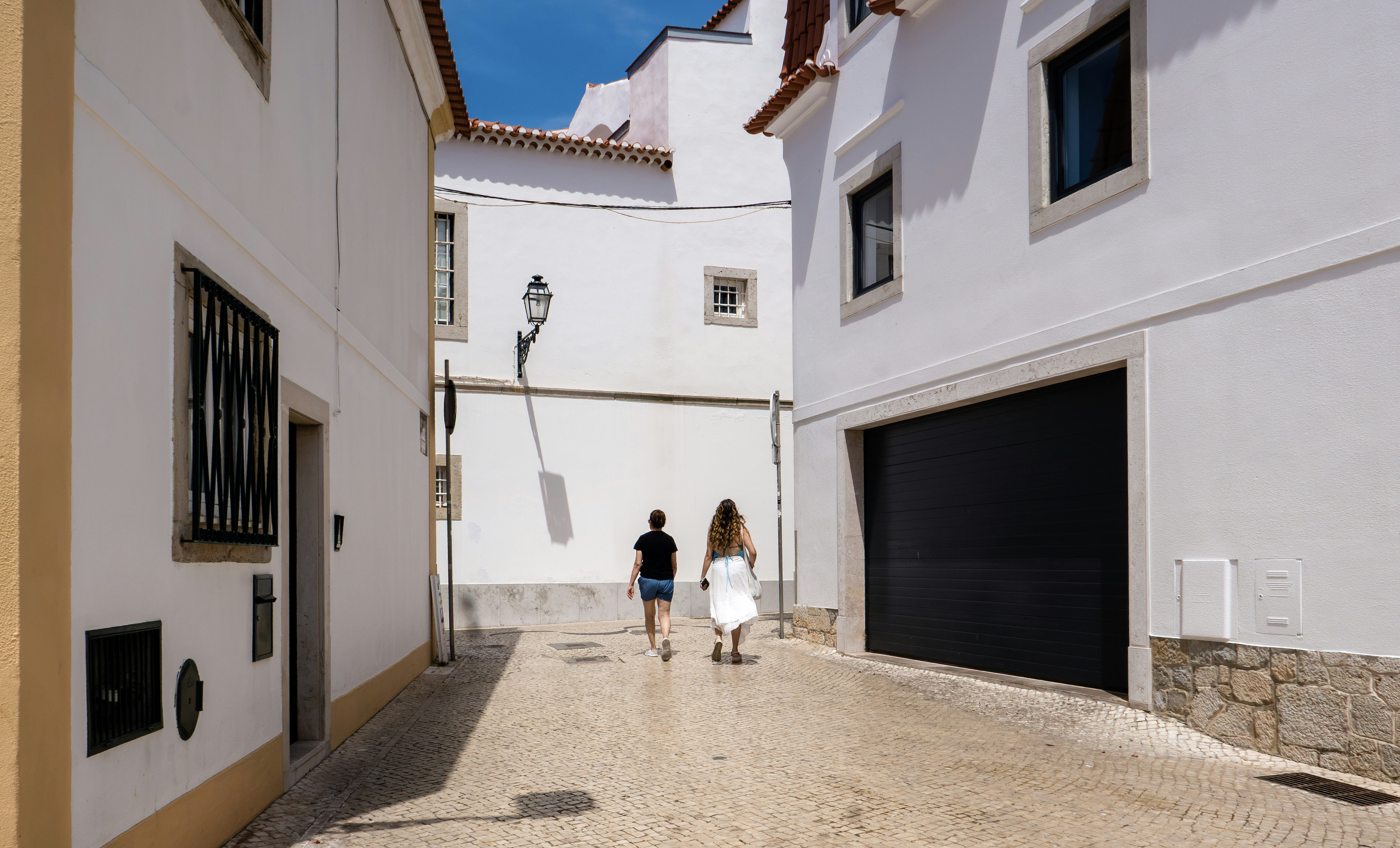Two people walk away from the camera down a sunlit, narrow white-walled street lined with simple buildings and a cobblestone path. A black garage door and a few windows add contrast to the scene.