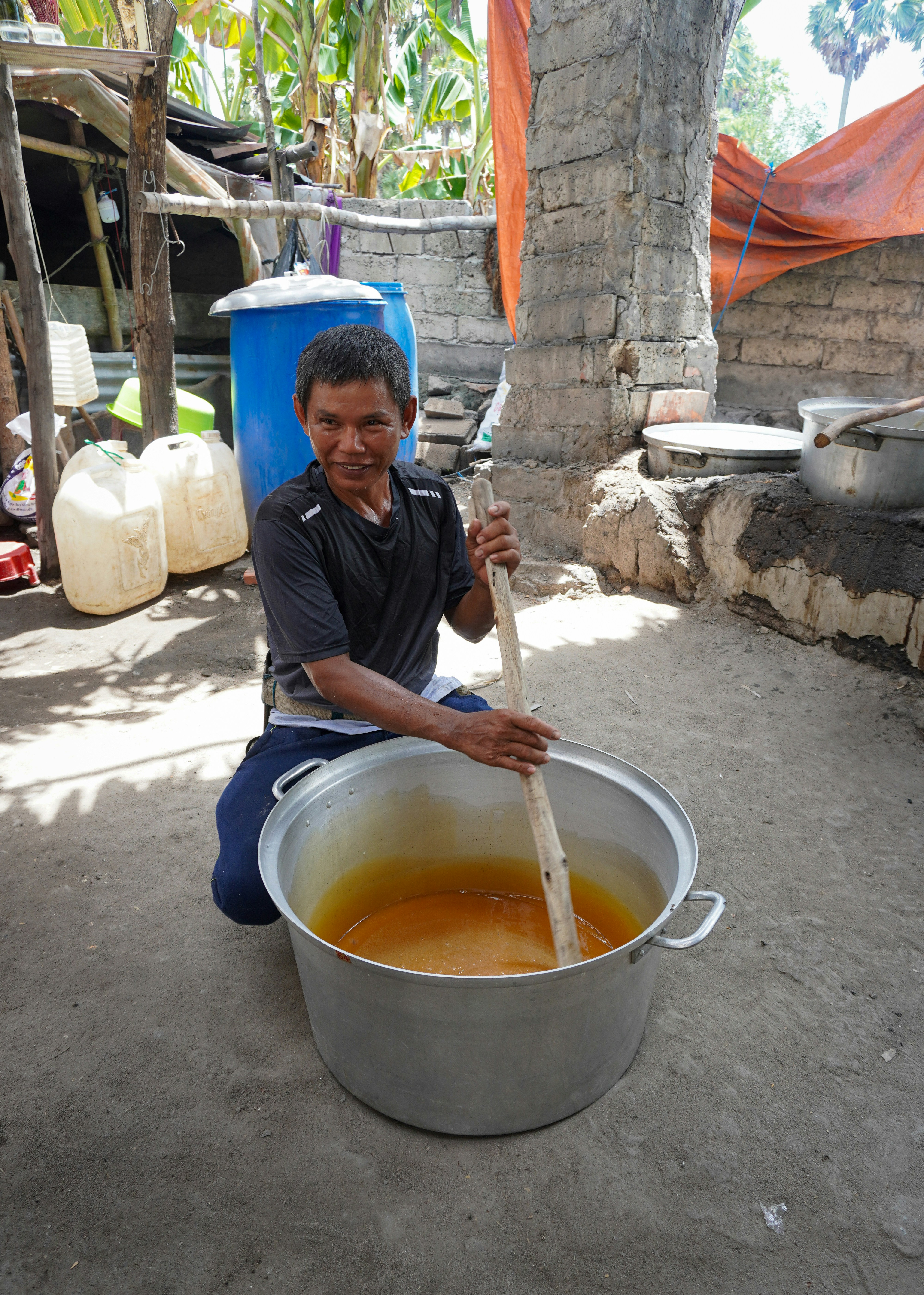 A man stirs a large pot outdoors in a sunlit yard, with colorful containers and a rustic backdrop framing the scene. The scene appears to capture a domestic cooking or liquid preparation moment.