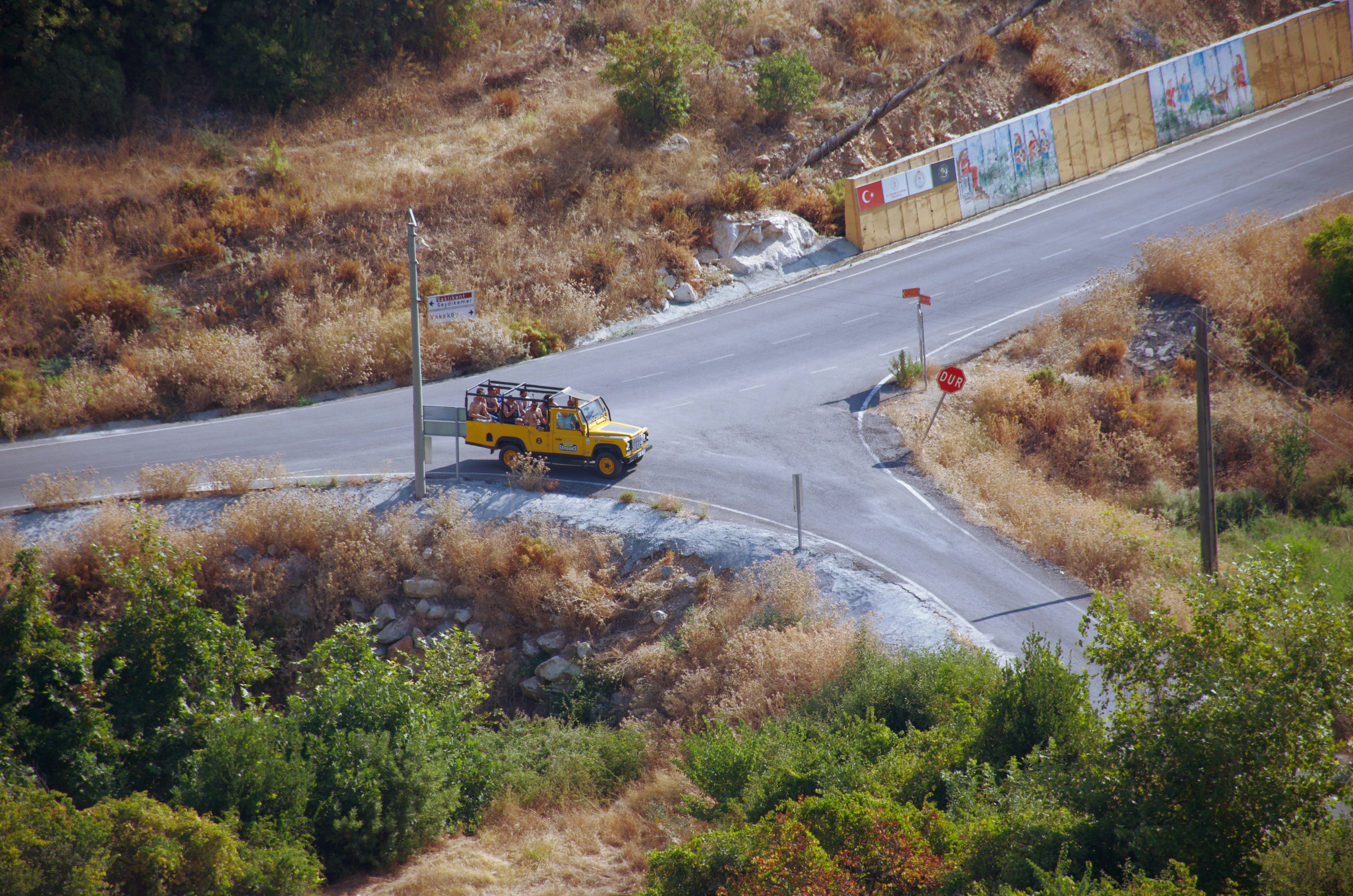 Yellow utility vehicle navigates a hairpin through a sunlit hillside road