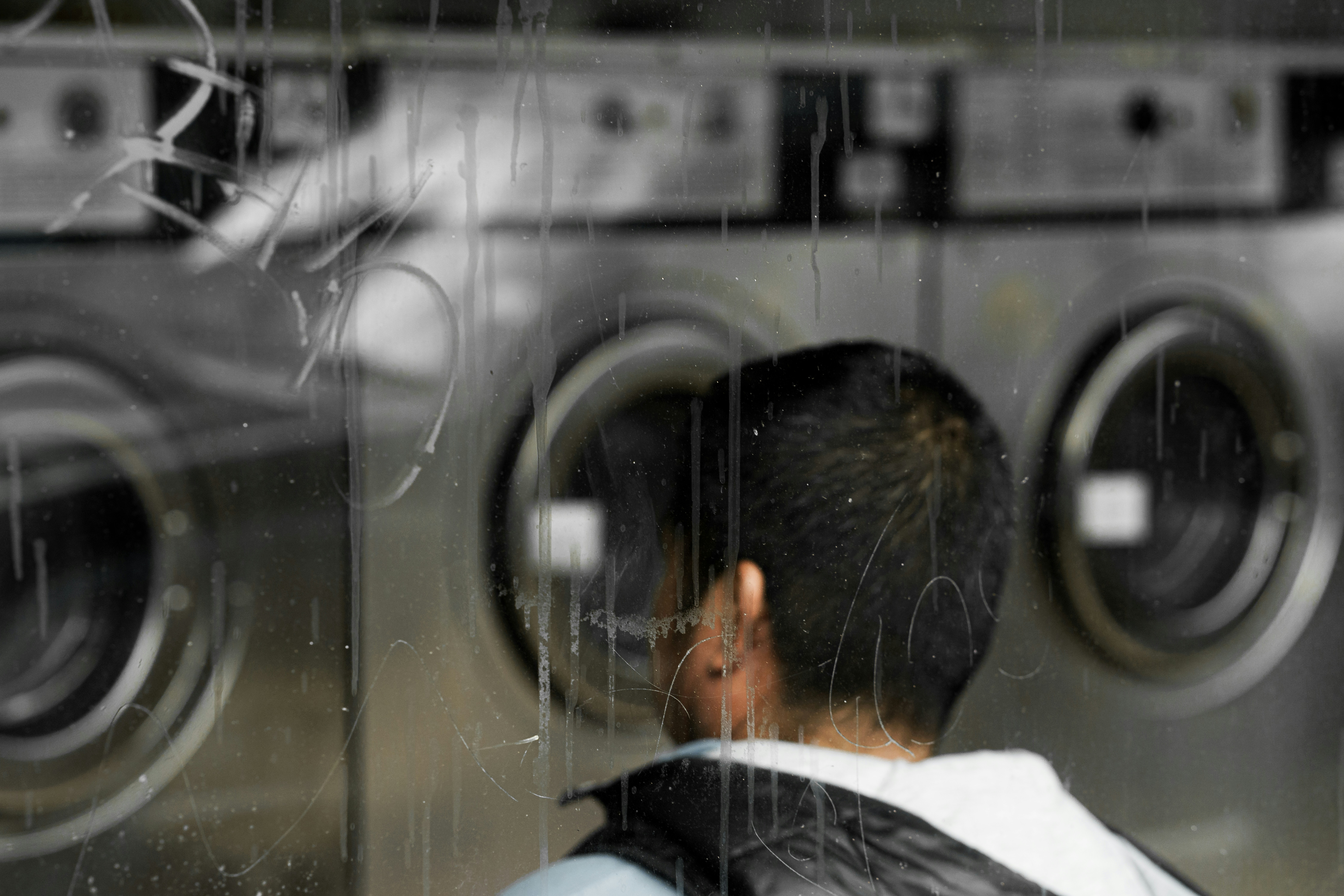 A man with a dark jacket stands with his back to the camera in front of a row of industrial washing machines; a reflective, slightly scratched glass creates a textured overlay, giving a candid street-like scene in a laundromat setting.