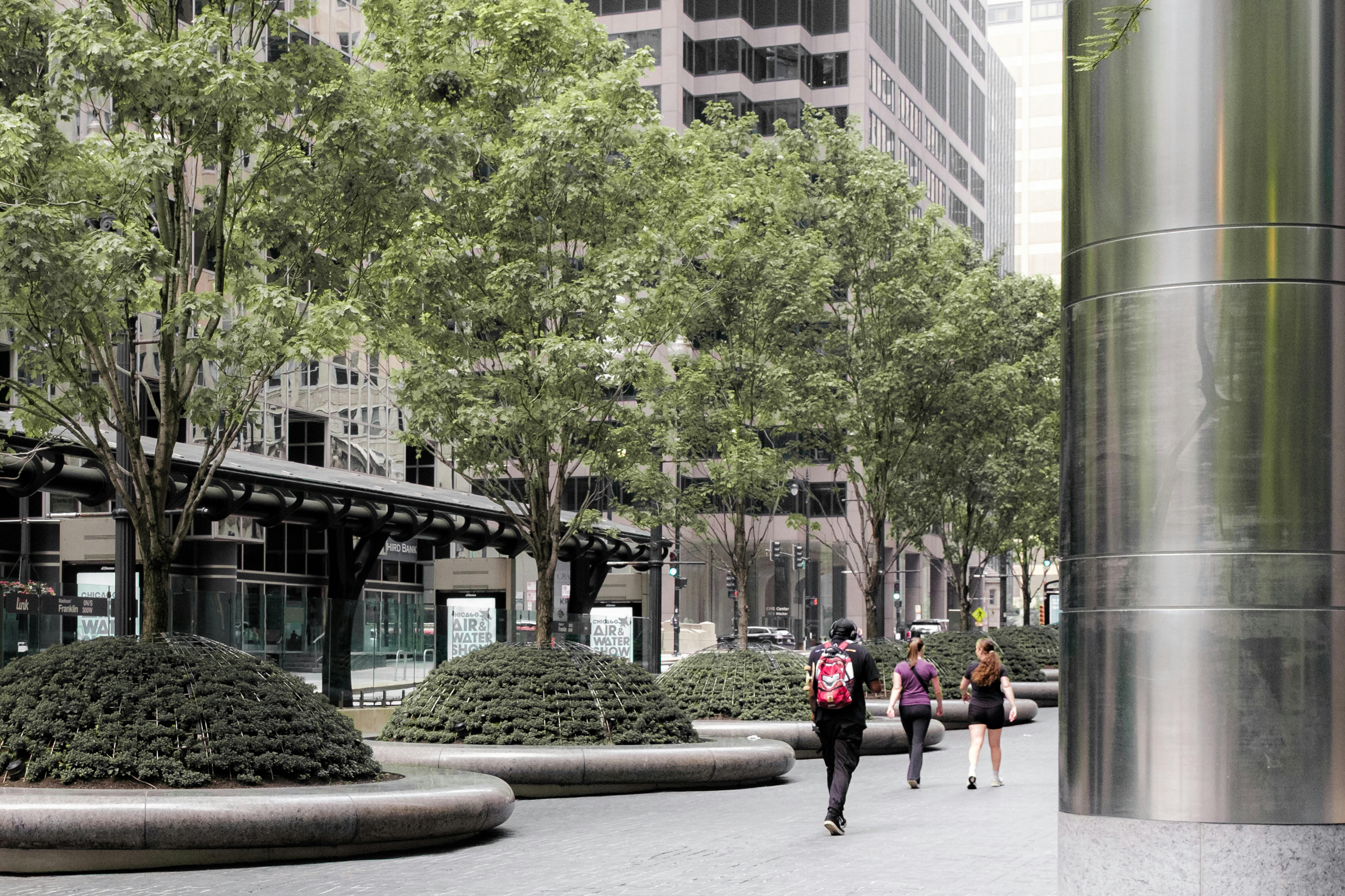 Urban Pace: Three pedestrians stride past a polished column in a sunlit plaza