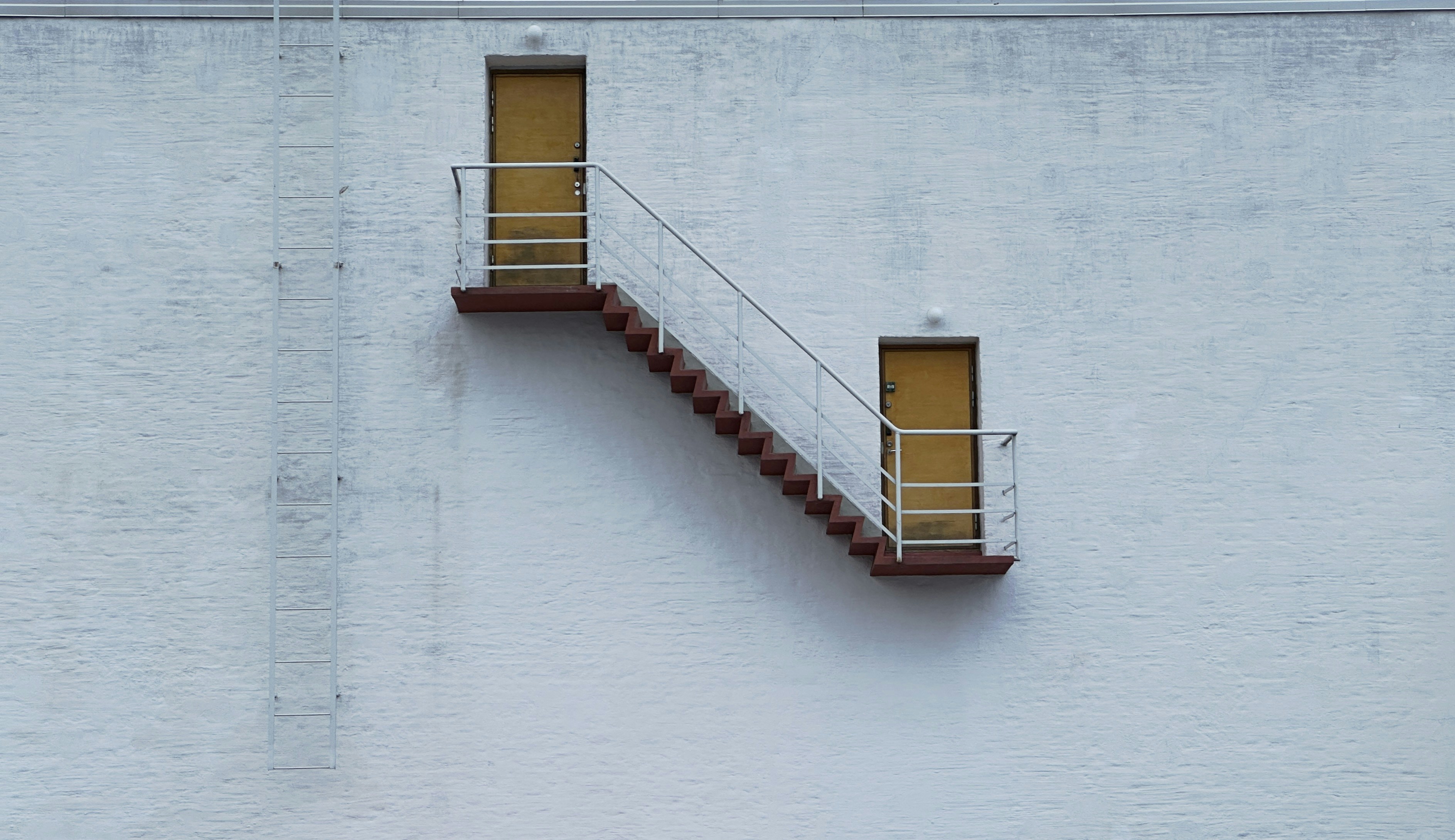 Minimalist scene of an exterior metal staircase cutting diagonally across a pale white wall, two yellow doors at each end; a quiet, architectural detail captured with soft lighting.