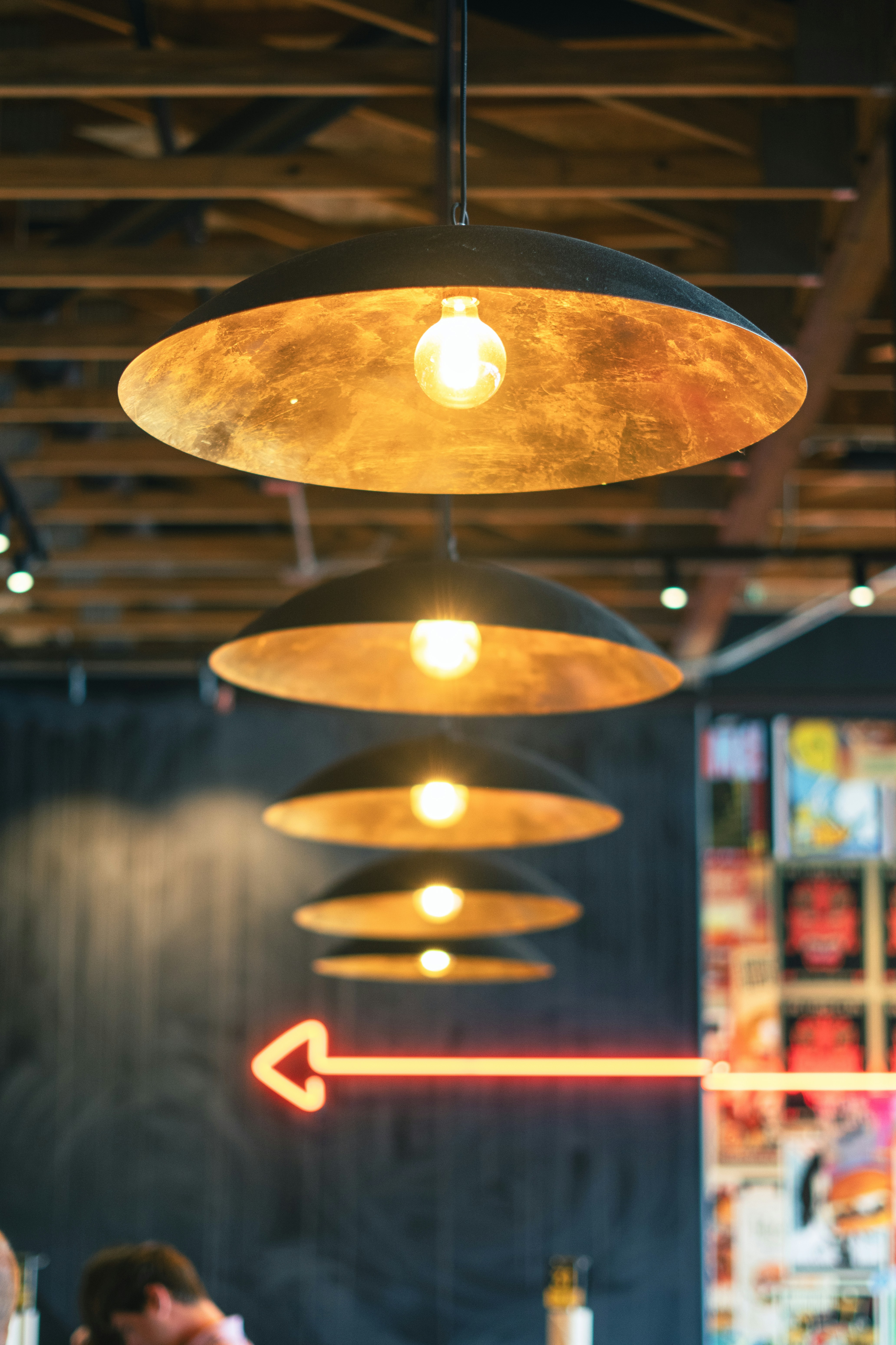 Copper pendant lights hang along a dimly lit ceiling with warm bulbs, casting a glow toward a backlit wall with posters and a neon arrow.