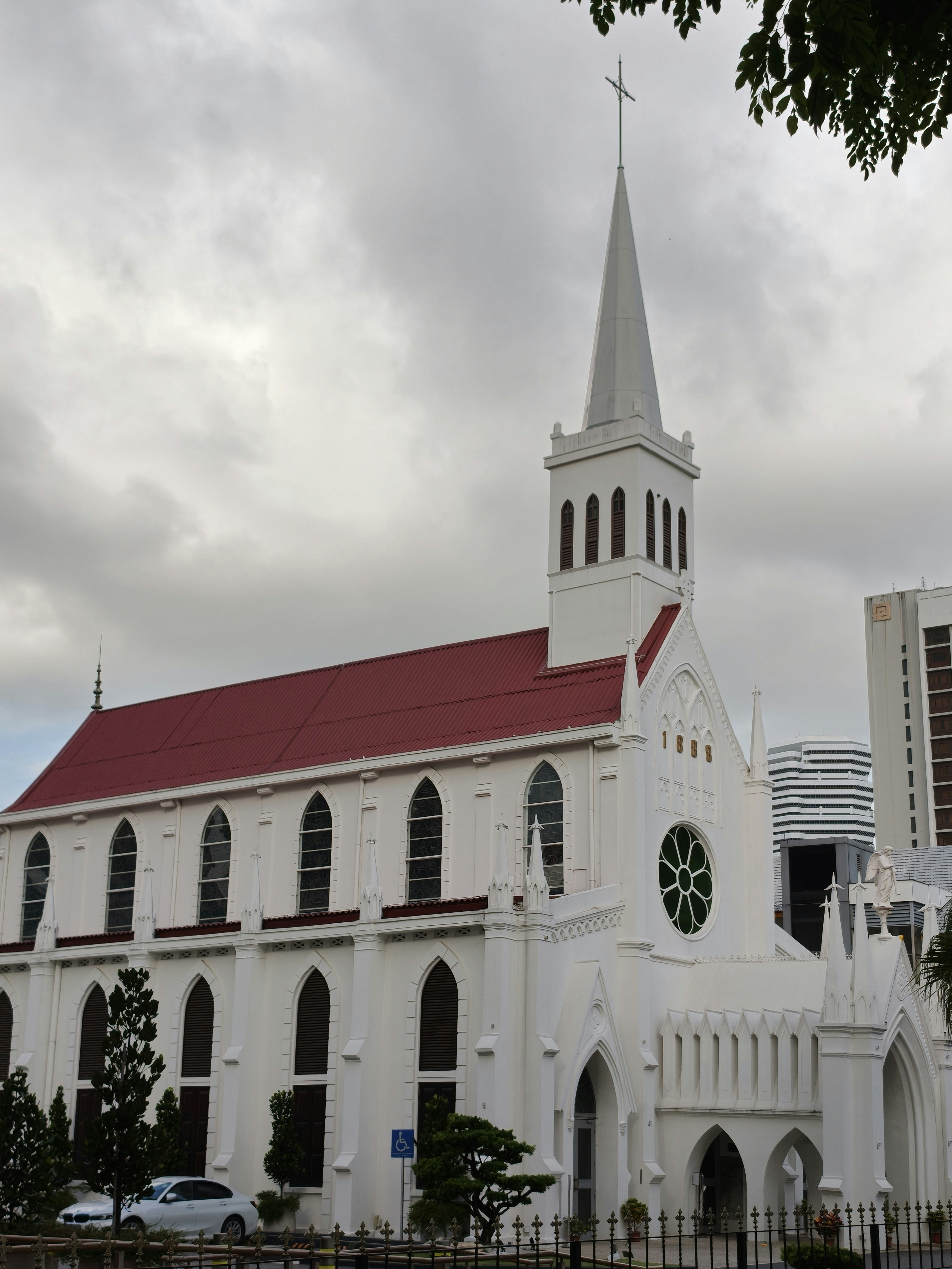 White church with red roof and tall steeple sits along a city street, featuring arched windows and a circular window, framed by overcast sky.