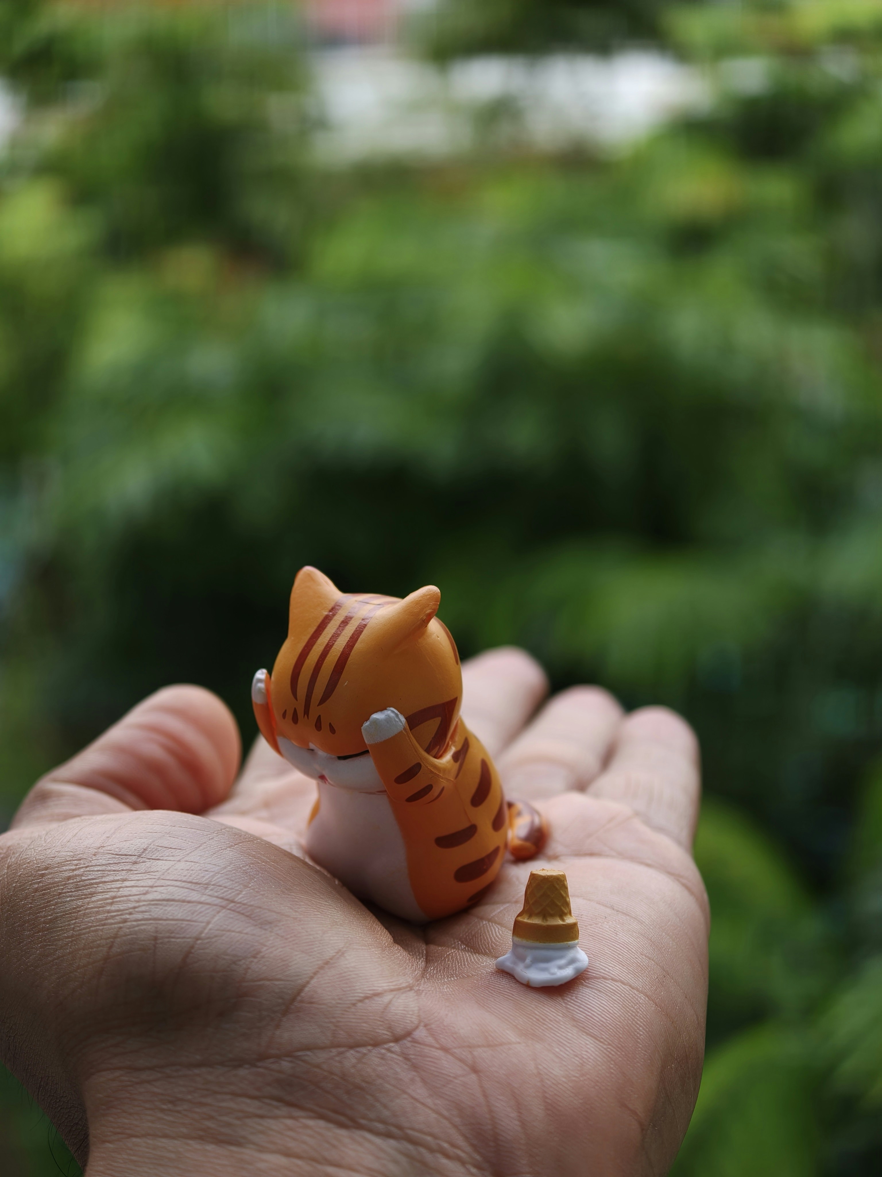 Small orange cat figurine resting on a palm with a tiny cone accessory, set against a blurred green background.