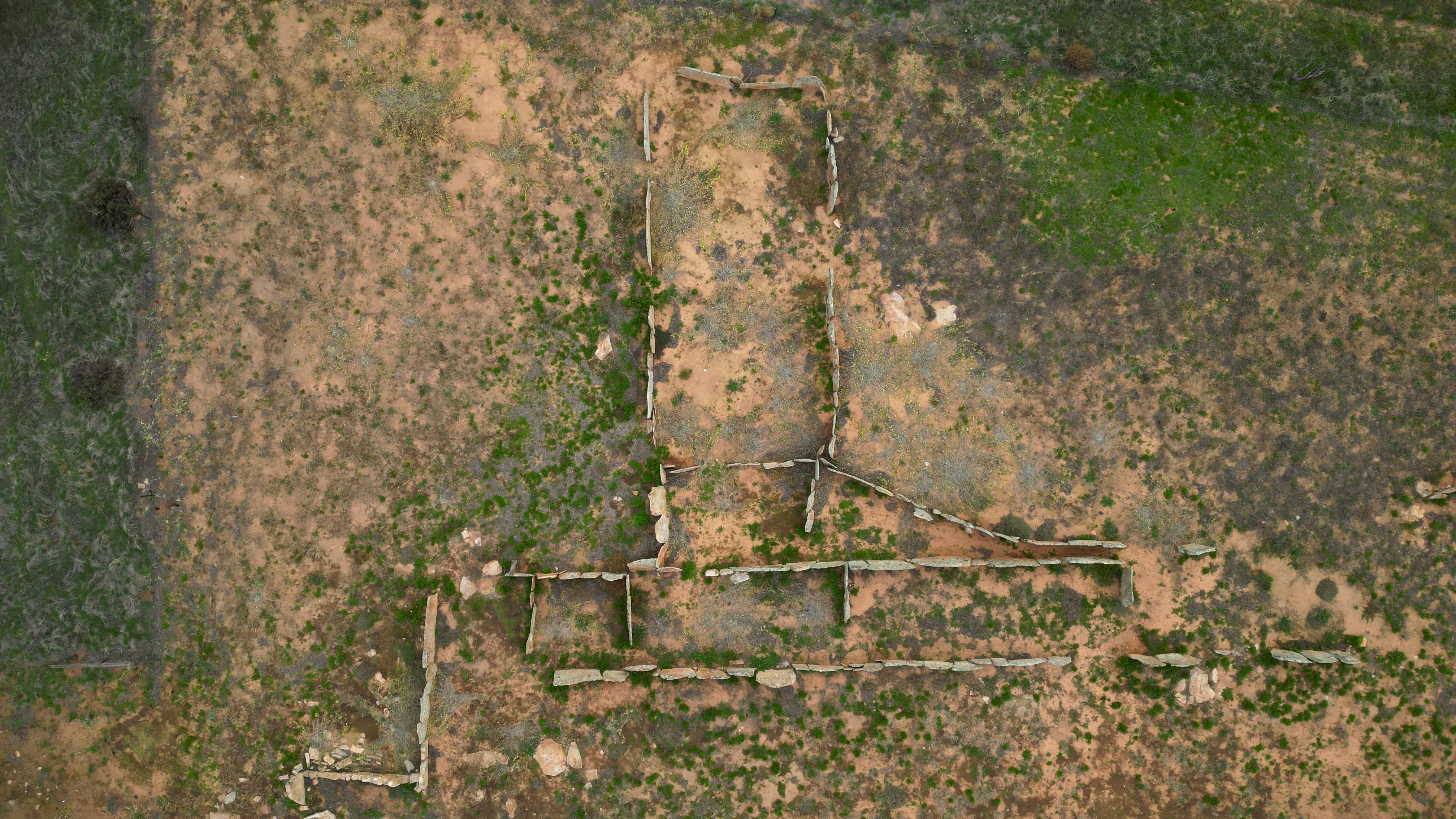 Mossy, weathered ruins spread across bare ground with patches of green; the central vertical remains hint at a former doorway or tower, suggesting an abandoned construct in a natural setting.