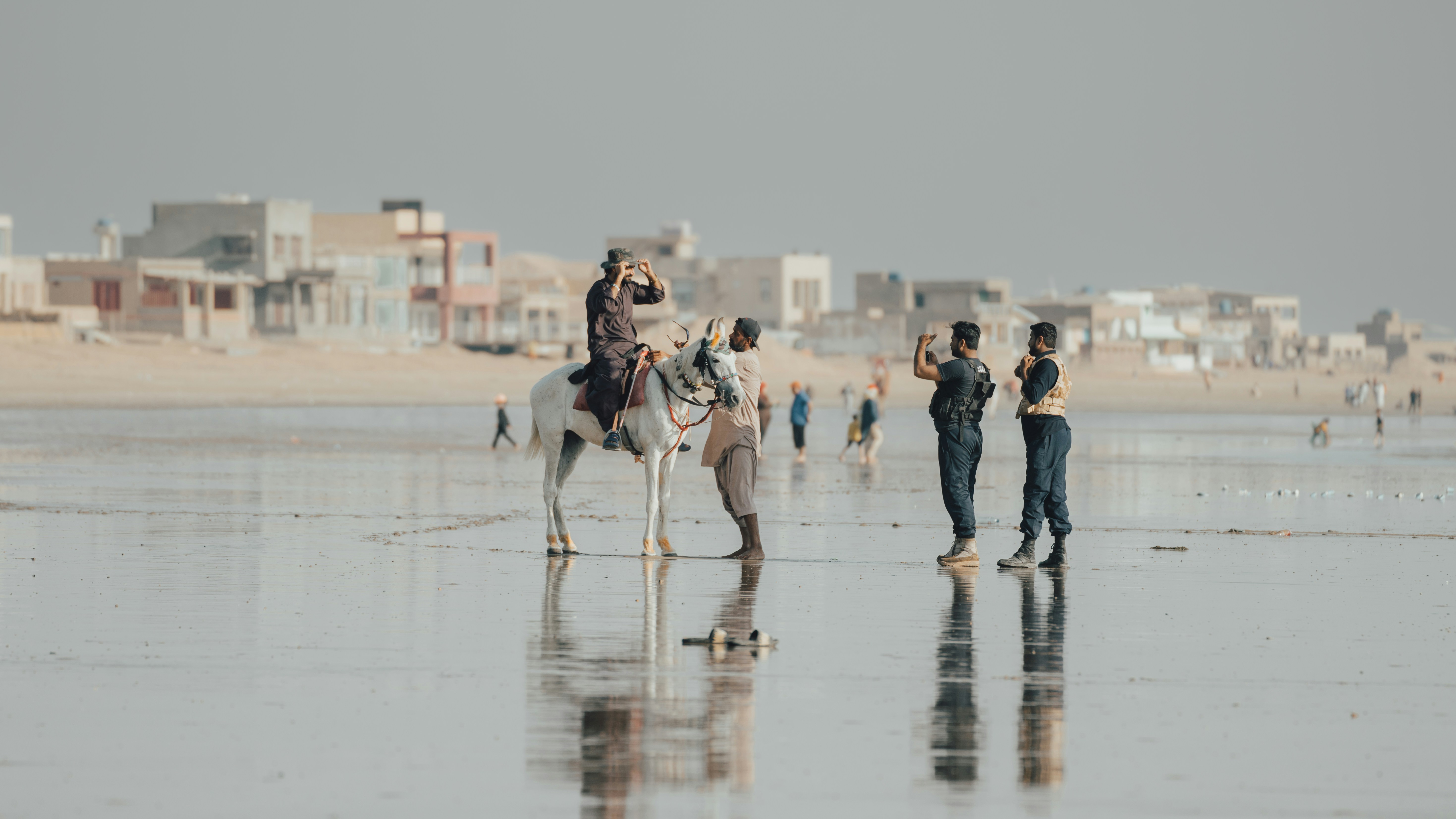 People photograph a rider on a white horse and others standing on a reflective shoreline with a distant town in the background.