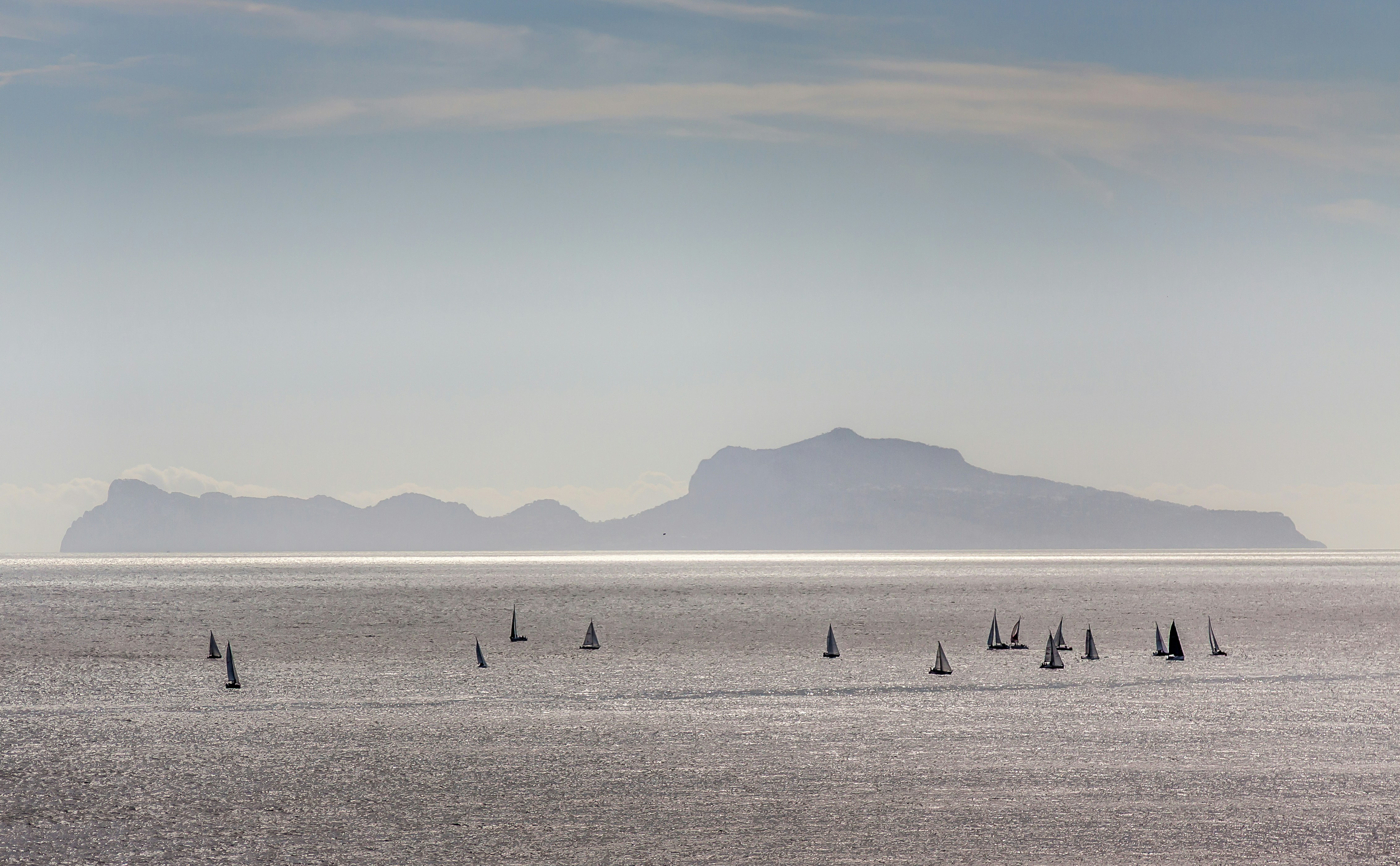 Low-angle view of a calm seascape with distant island silhouette and scattered sailboats on a reflective tidal plain.