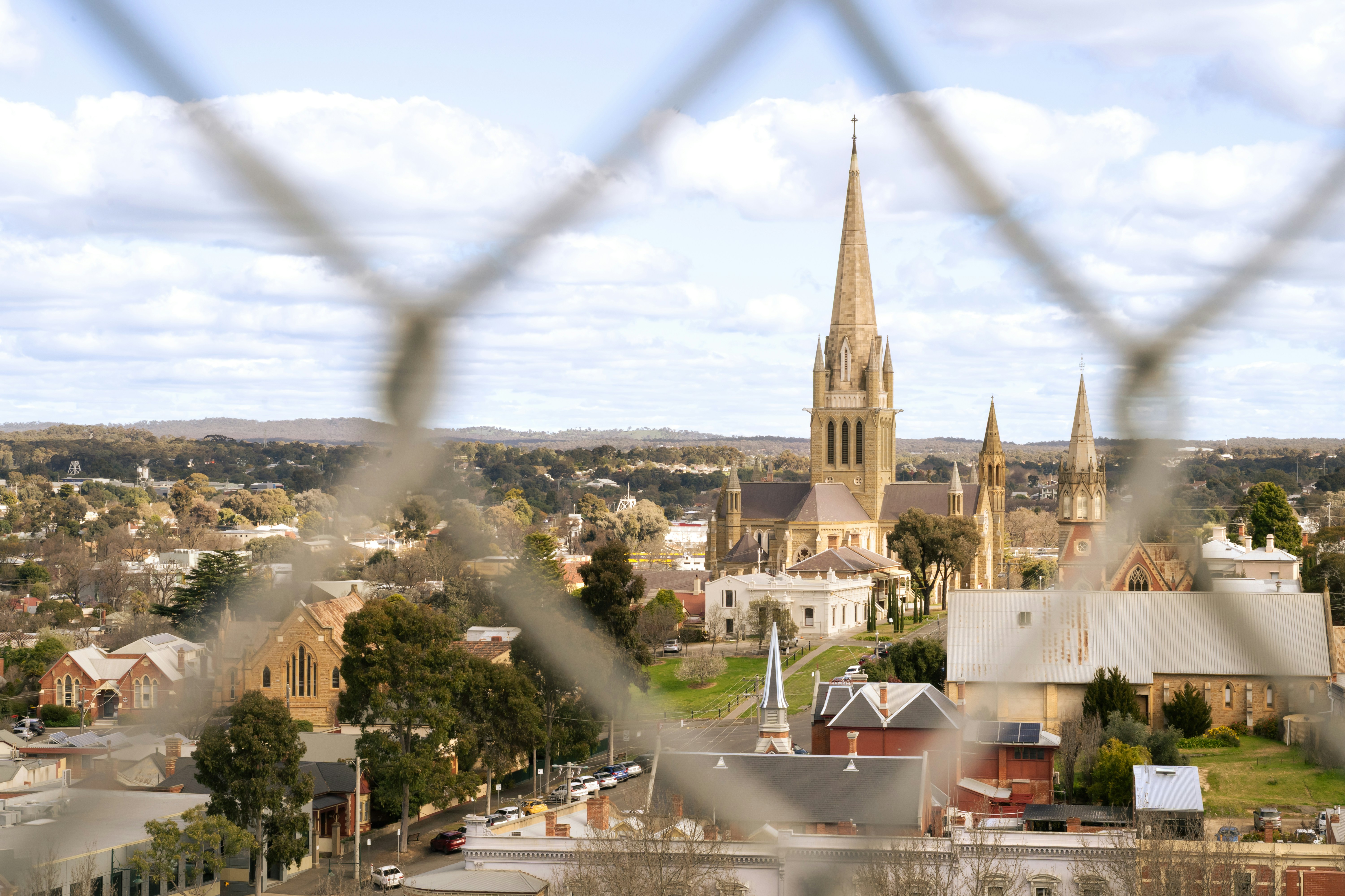 Historic church towers rise beyond a small town, framed by a blurred chain-link fence in the foreground; a quiet civicscape beneath a bright, slightly overcast sky.