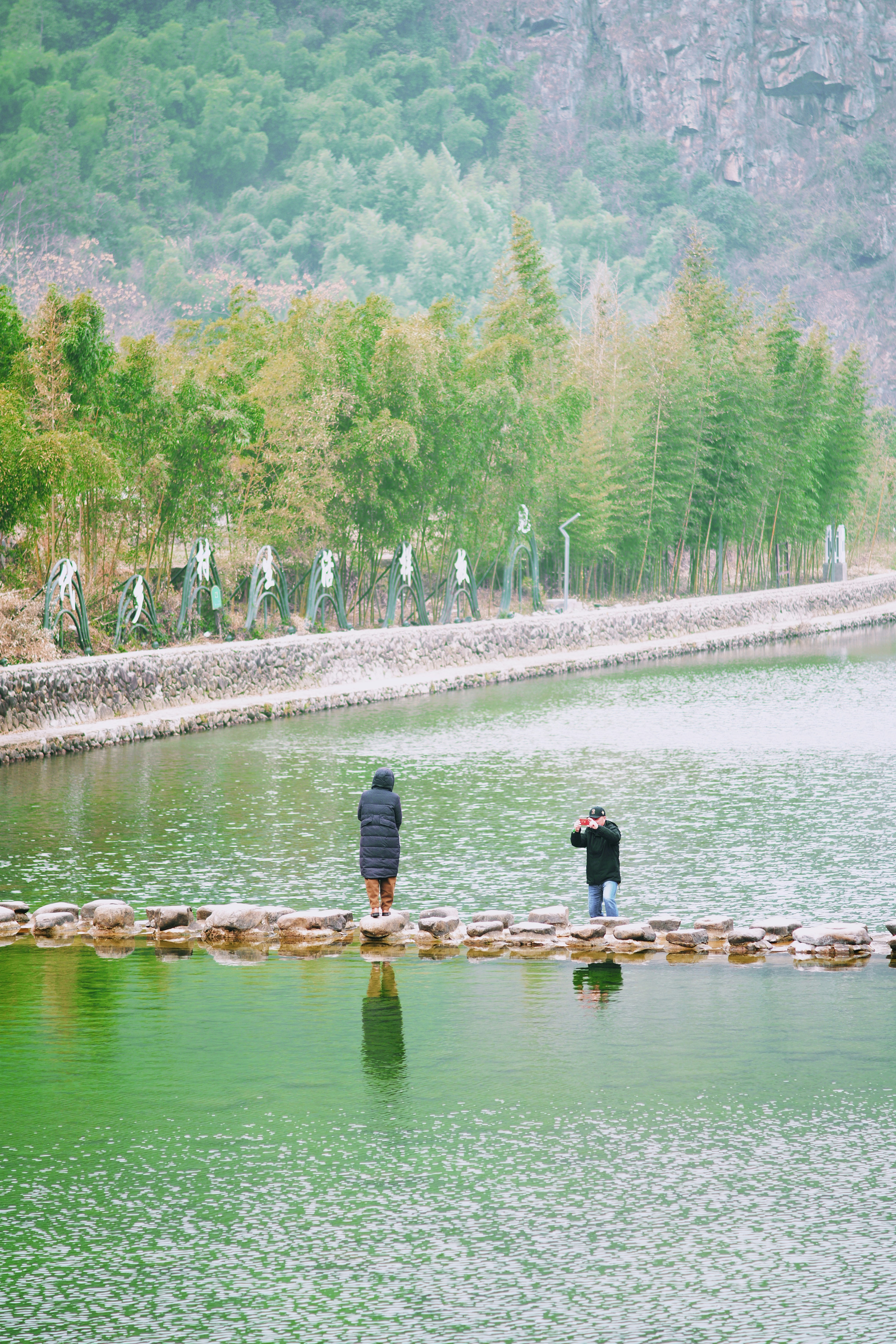 Two figures on a stone bridge over a calm green river with a forested backdrop