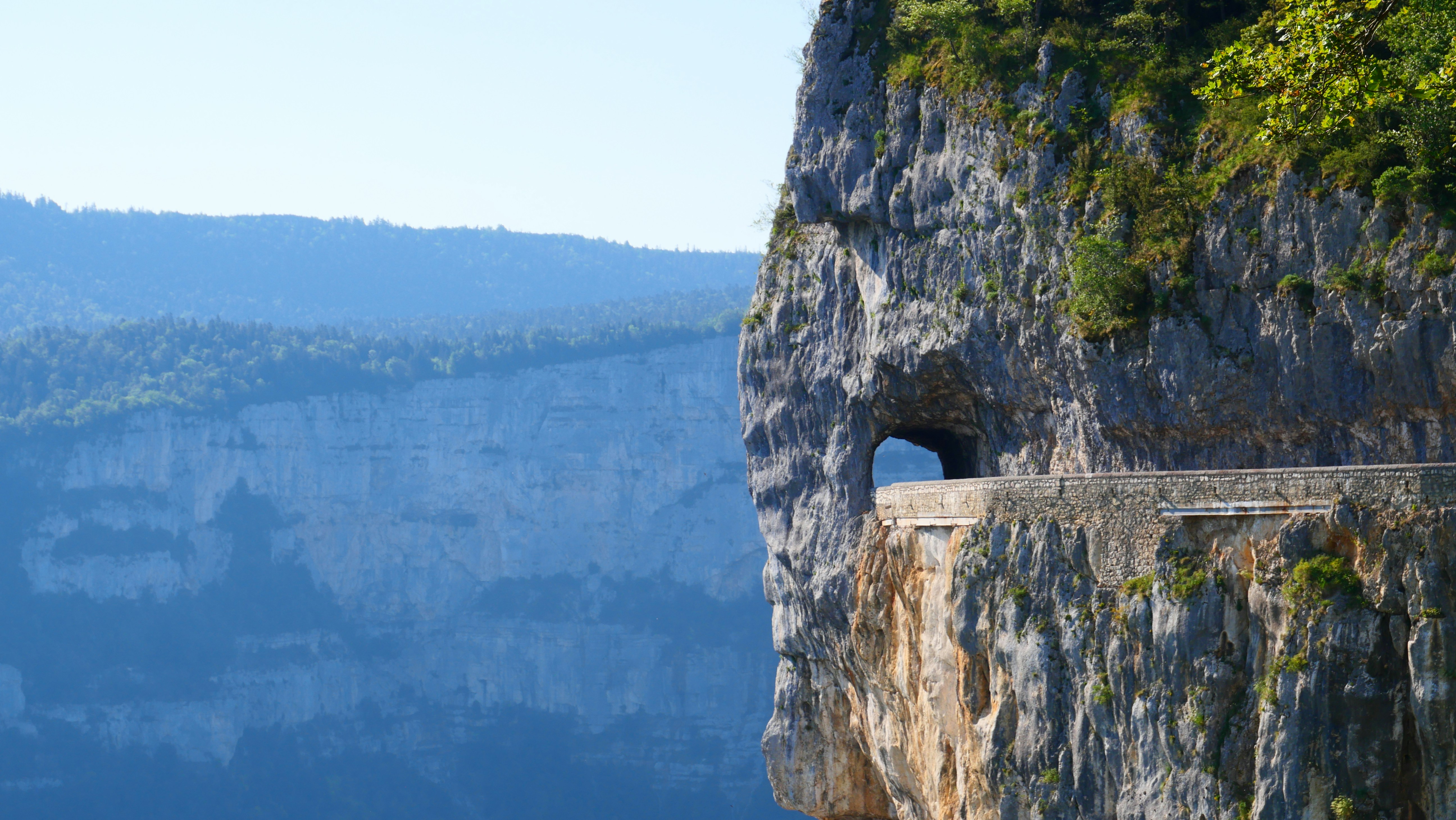 A rugged cliffside road with a small tunnel carved into rock spans a sheer drop, carved guardwall along a narrow bridge over layered canyon walls under clear daylight.