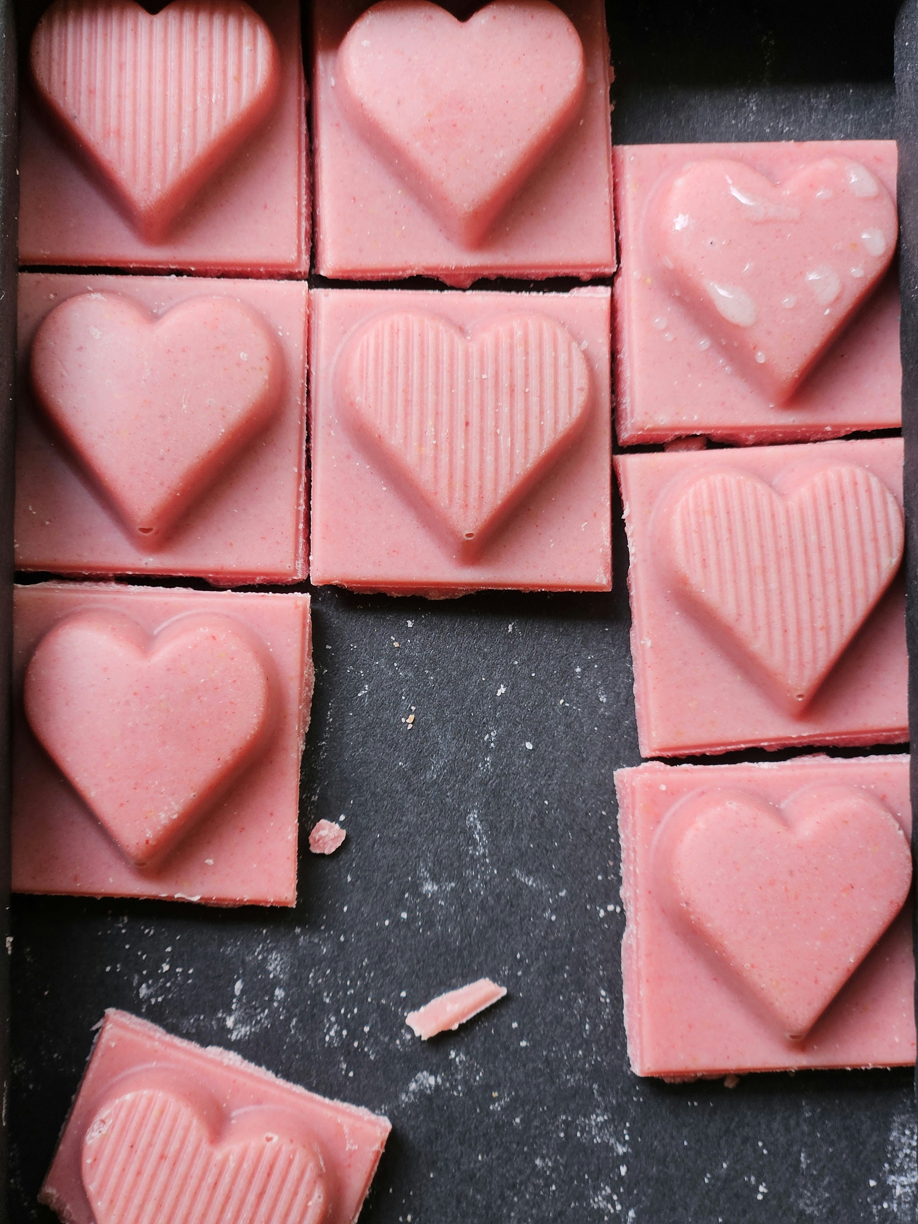 Close-up of pink heart-shaped chocolate squares aligned in a neat grid on a dark surface. The focus highlights texture and subtle gloss, with a few broken pieces nearby.