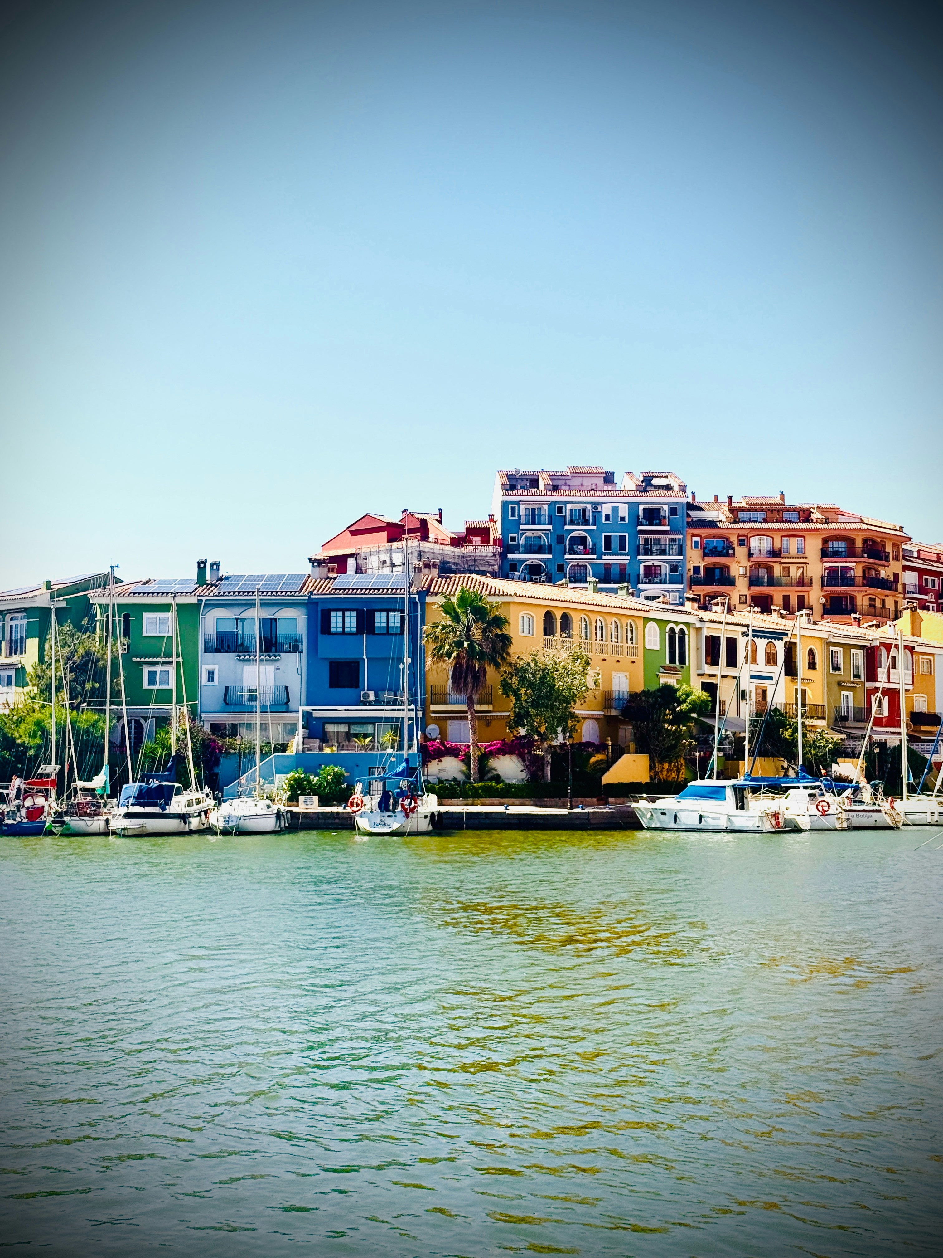Colorful row houses along a marina with boats, reflected water, and a clear blue sky.