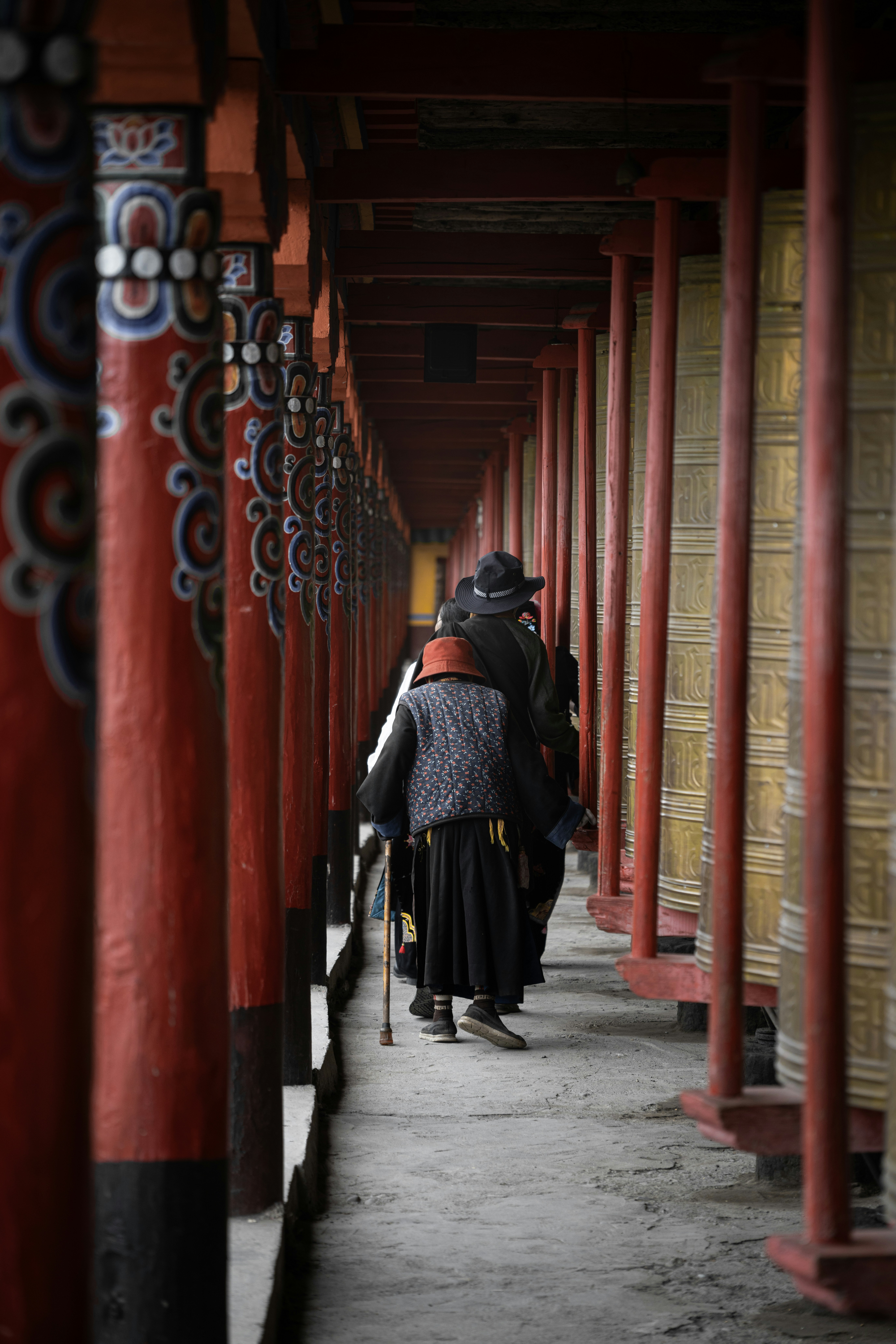 Two visitors walk away through a narrow red-columned corridor of a traditional temple, with patterned pillars and ornate walls guiding toward a distant doorway.