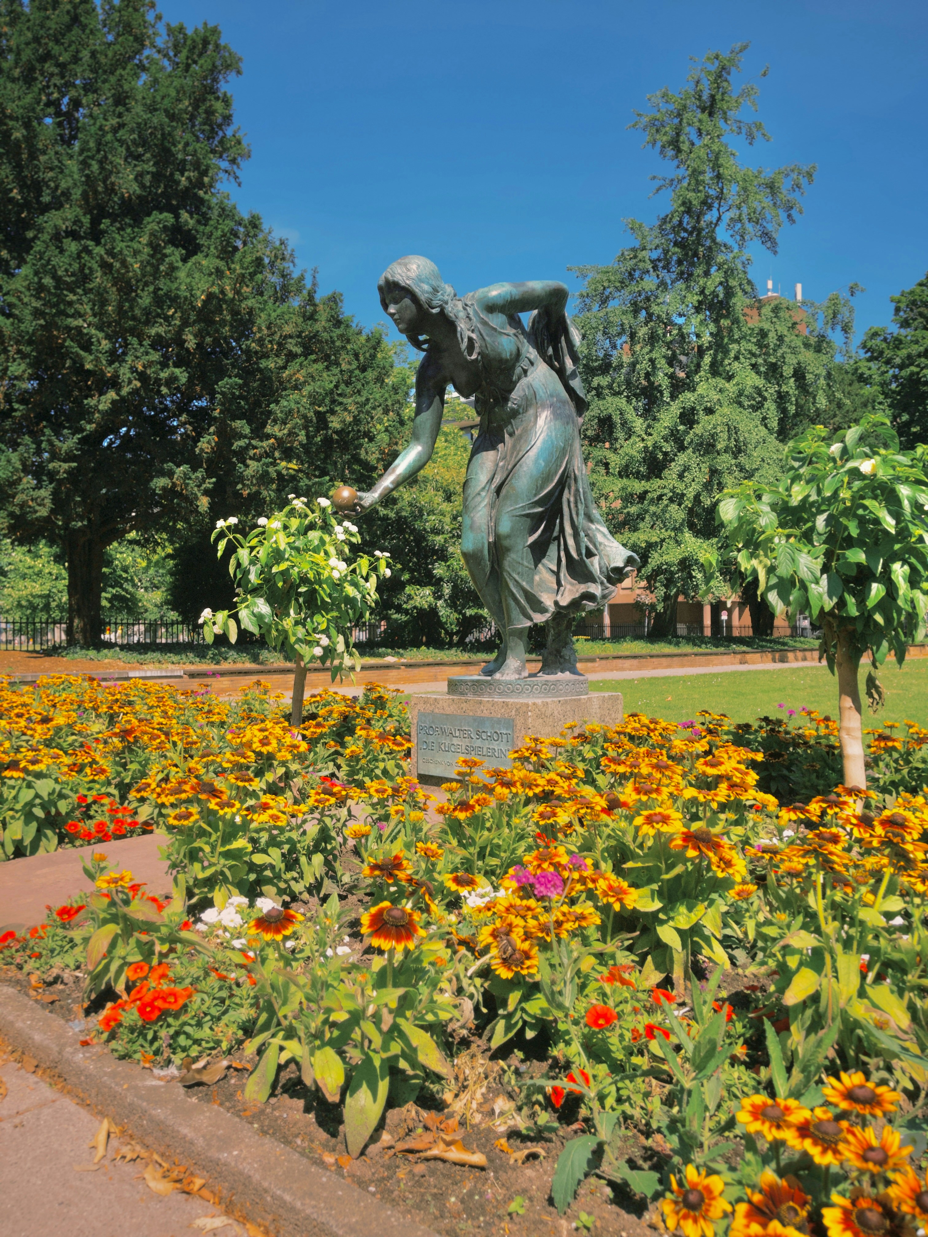 Bronze statue of a graceful woman reaching toward a small tree, surrounded by a vibrant bed of marigolds and framed by a sunny park setting.
