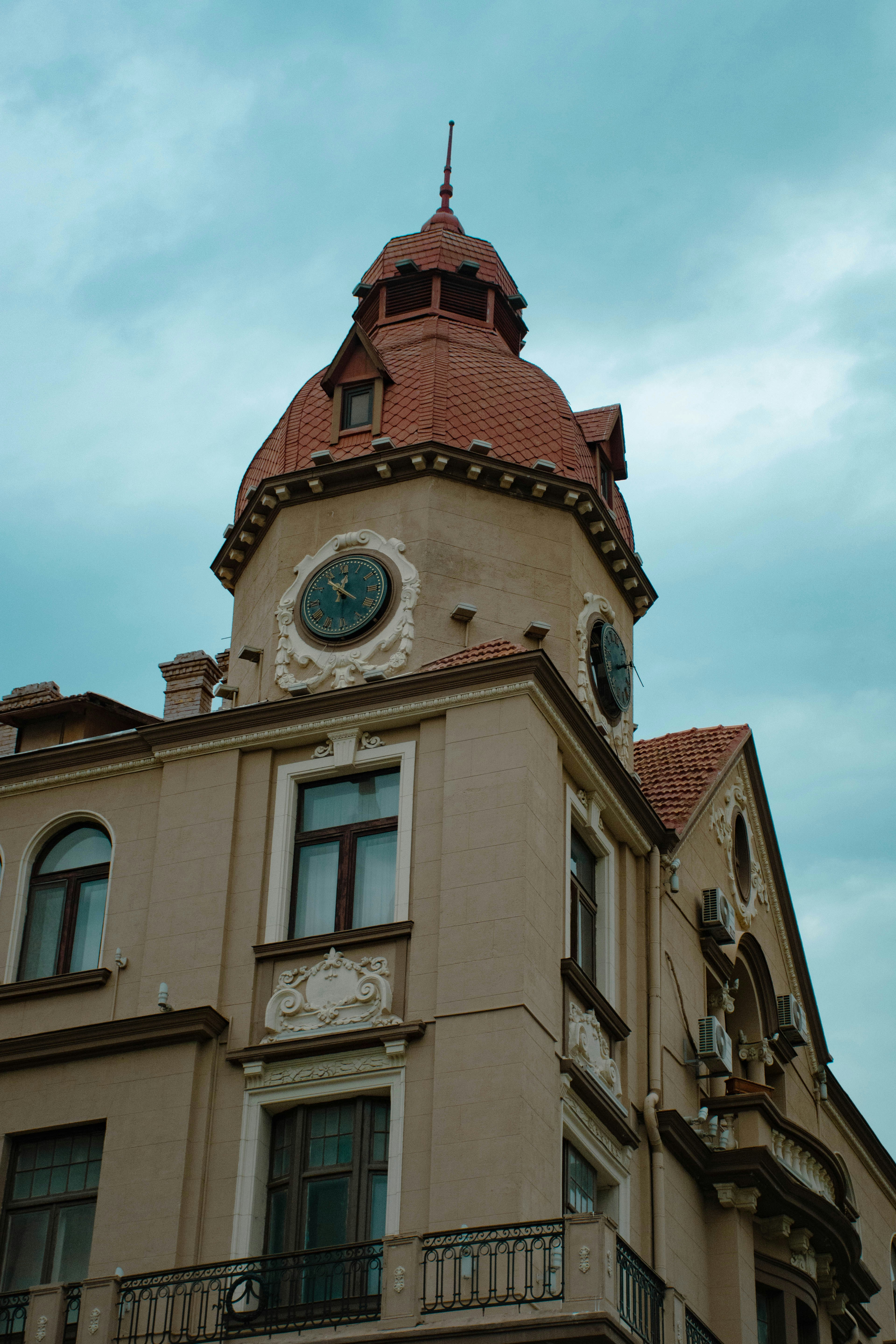 Elegant city clocktower rises from a beige ornate building against a pale blue sky. Architectural details and balconies sit beneath a red-tiled dome, suggesting a historic urban scene.