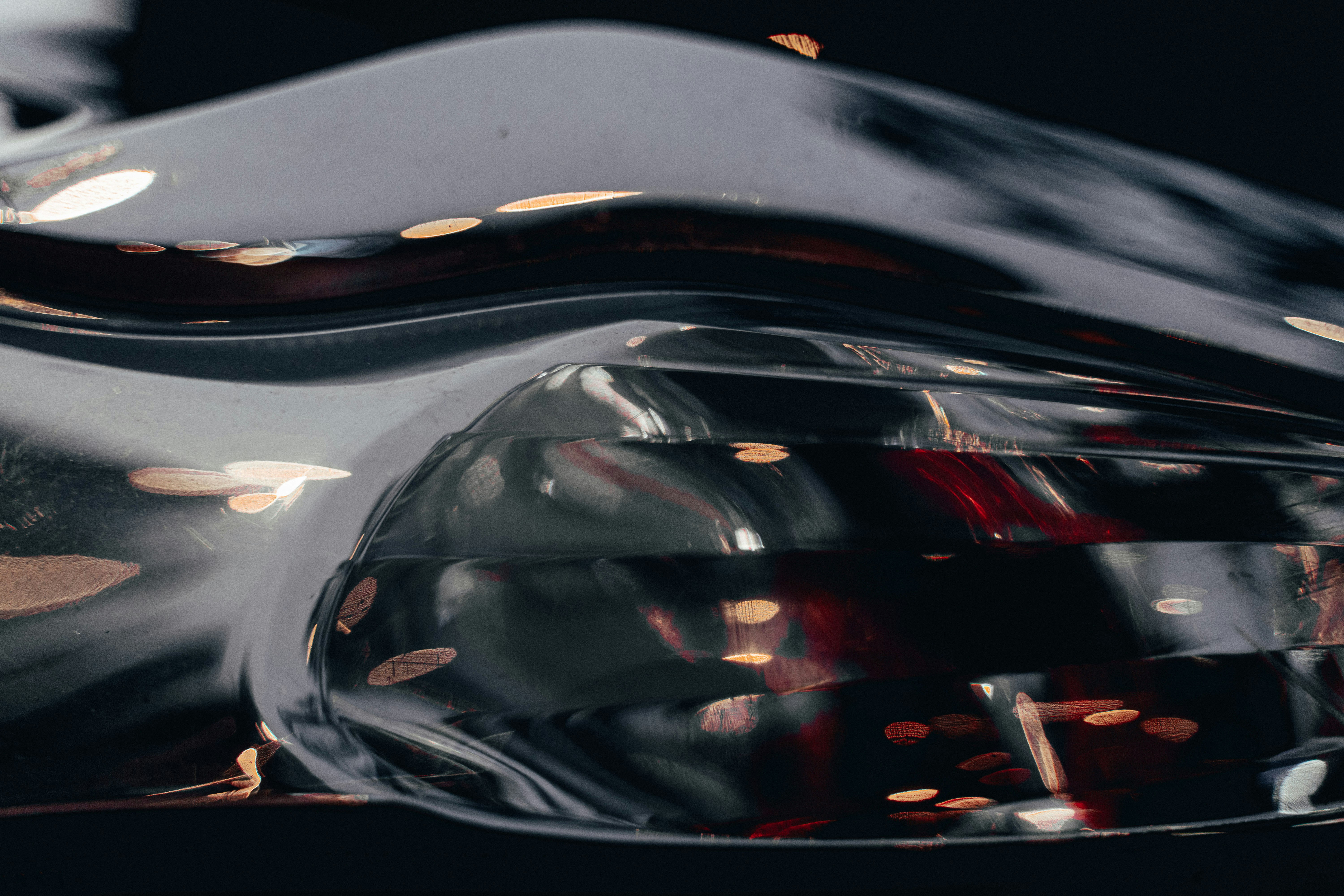 Glossy, abstract metal surface with blurred light reflections forming orange highlights and dark contours. A close-up of liquid-like curves suggests a sculpture or automotive surface detail.