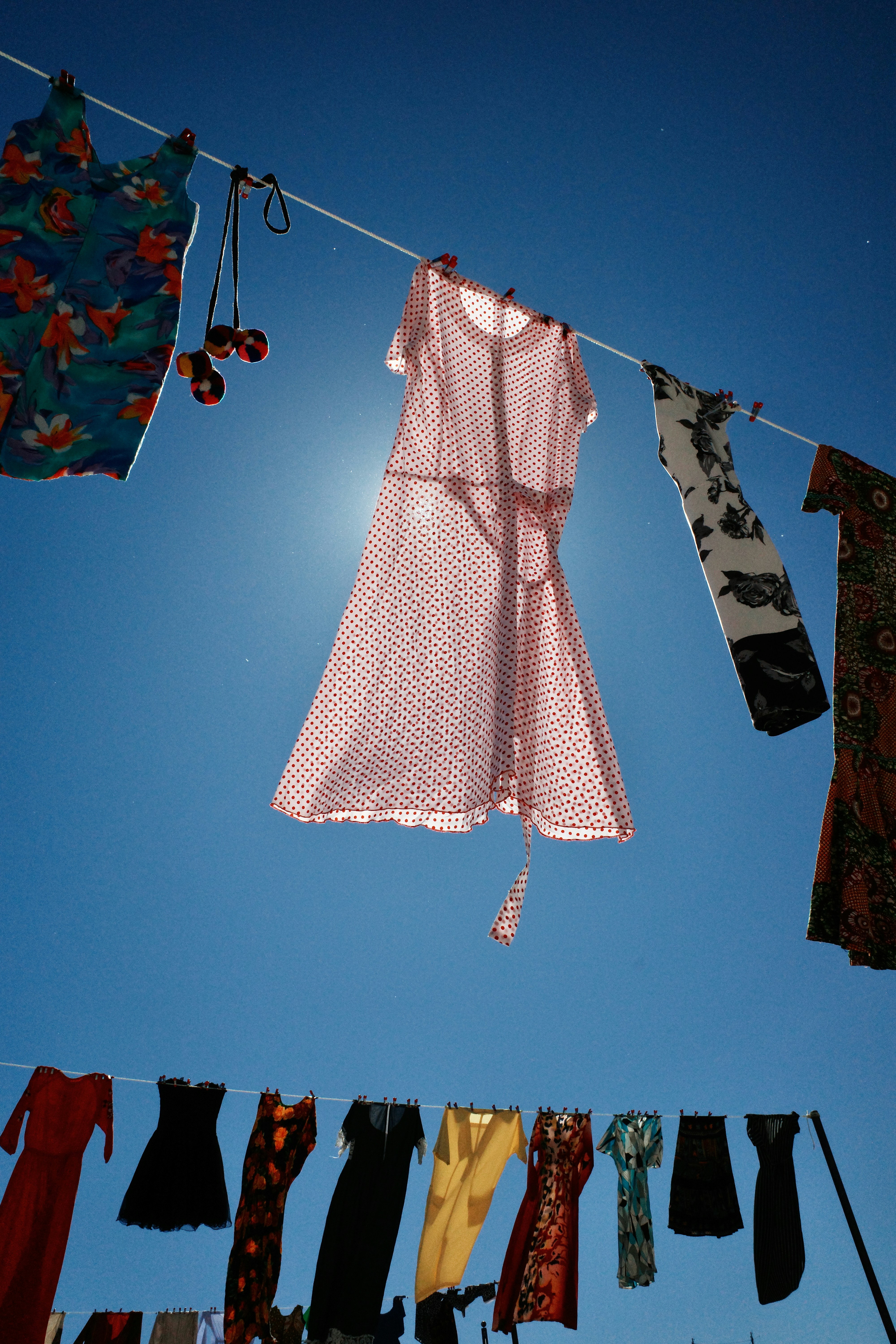 A bright pink polka-dot dress hangs on a clothesline against a clear blue sky, with other colorful garments fluttering along the line below.