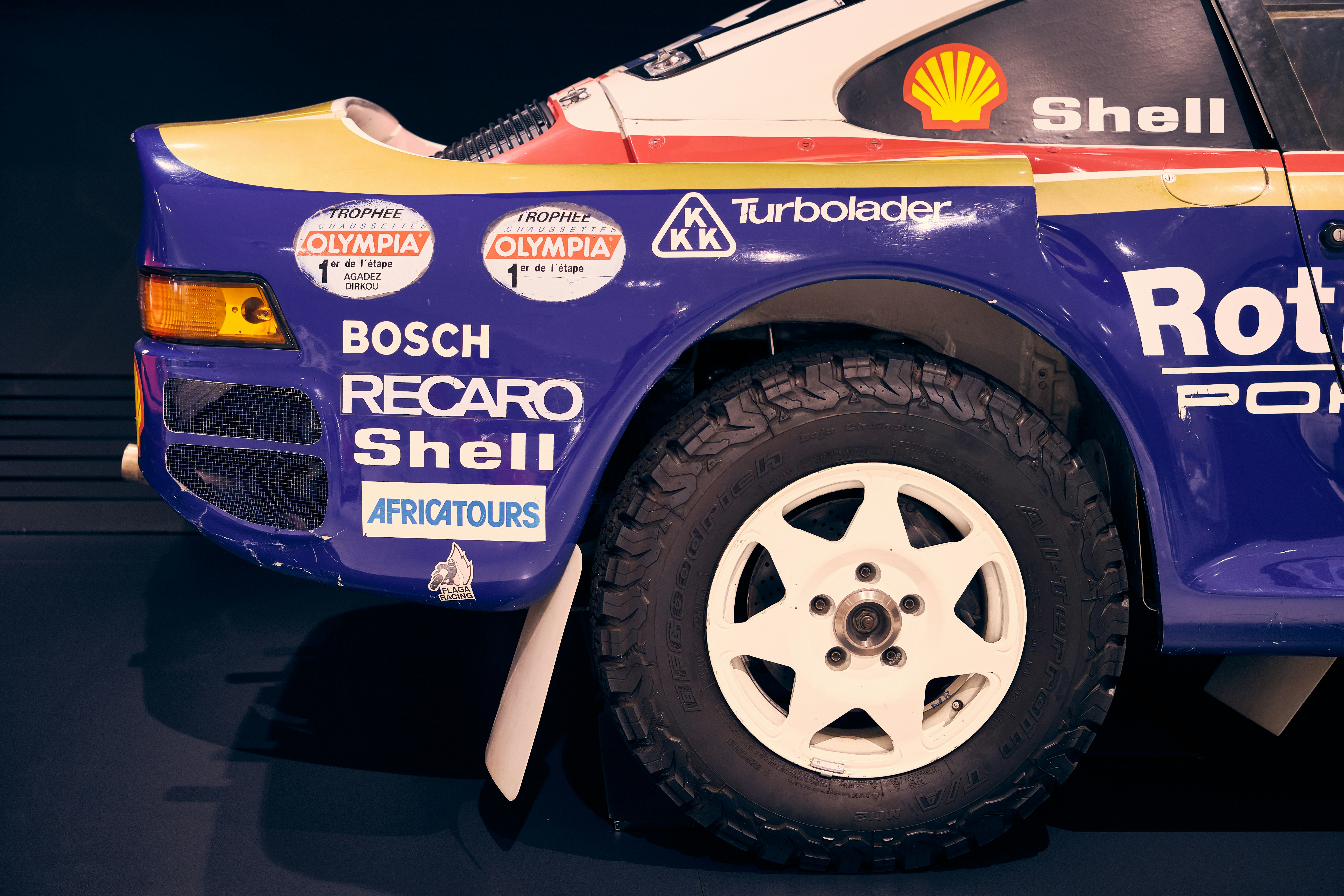 Close-up of a rally car front fender and wheel with sponsor decals in a vivid blue and purple livery.