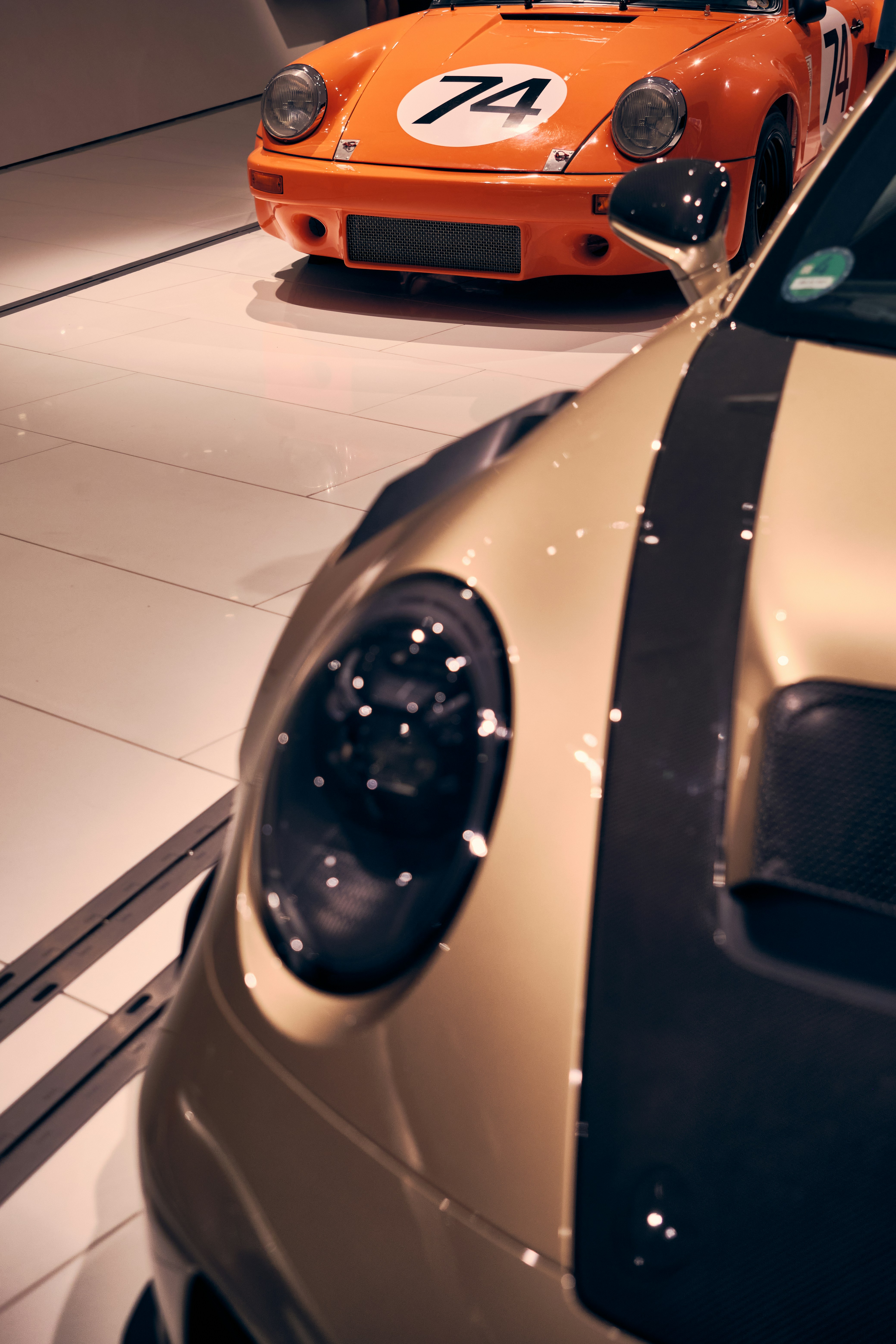 Orange racer emerges from the show floor, peeking behind a tan Porsche