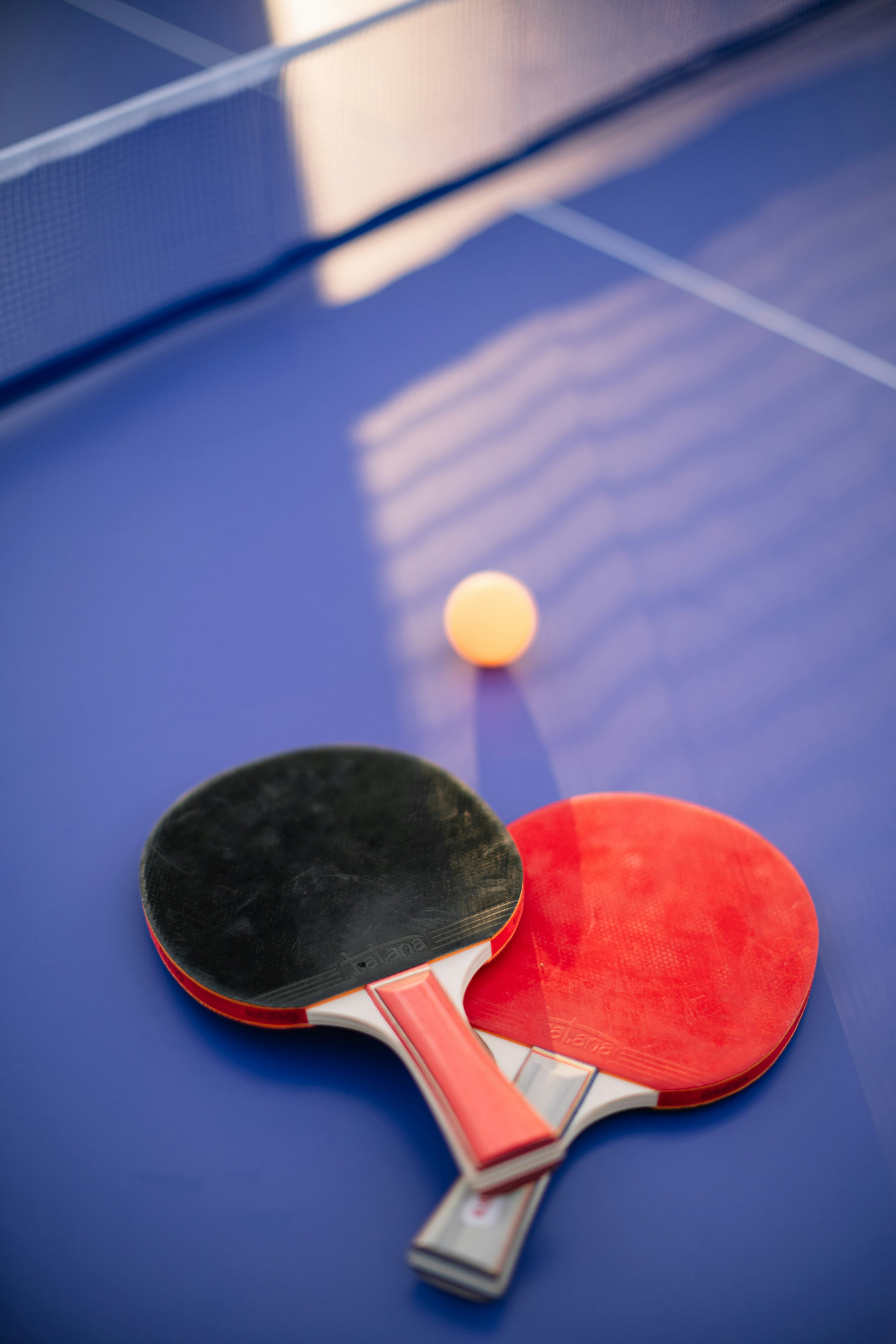 Two table tennis paddles lie on a blue table with a small orange ball resting between them, a shallow depth of field emphasizing the rackets while soft reflections imprint across the surface.