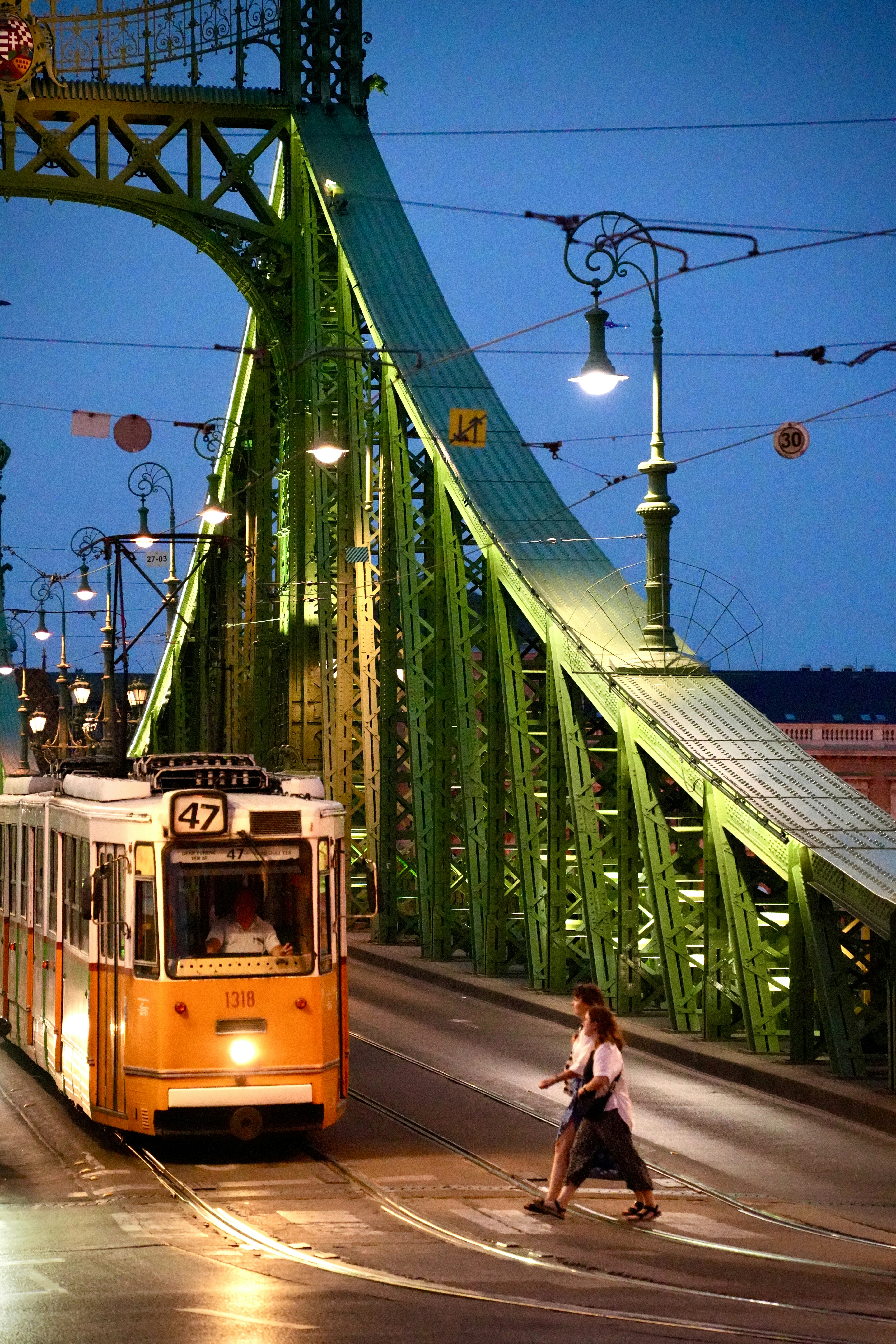 A yellow tram travels along tracks on a green metal suspension bridge at dusk, as pedestrians cross the road beneath glowing street lamps.