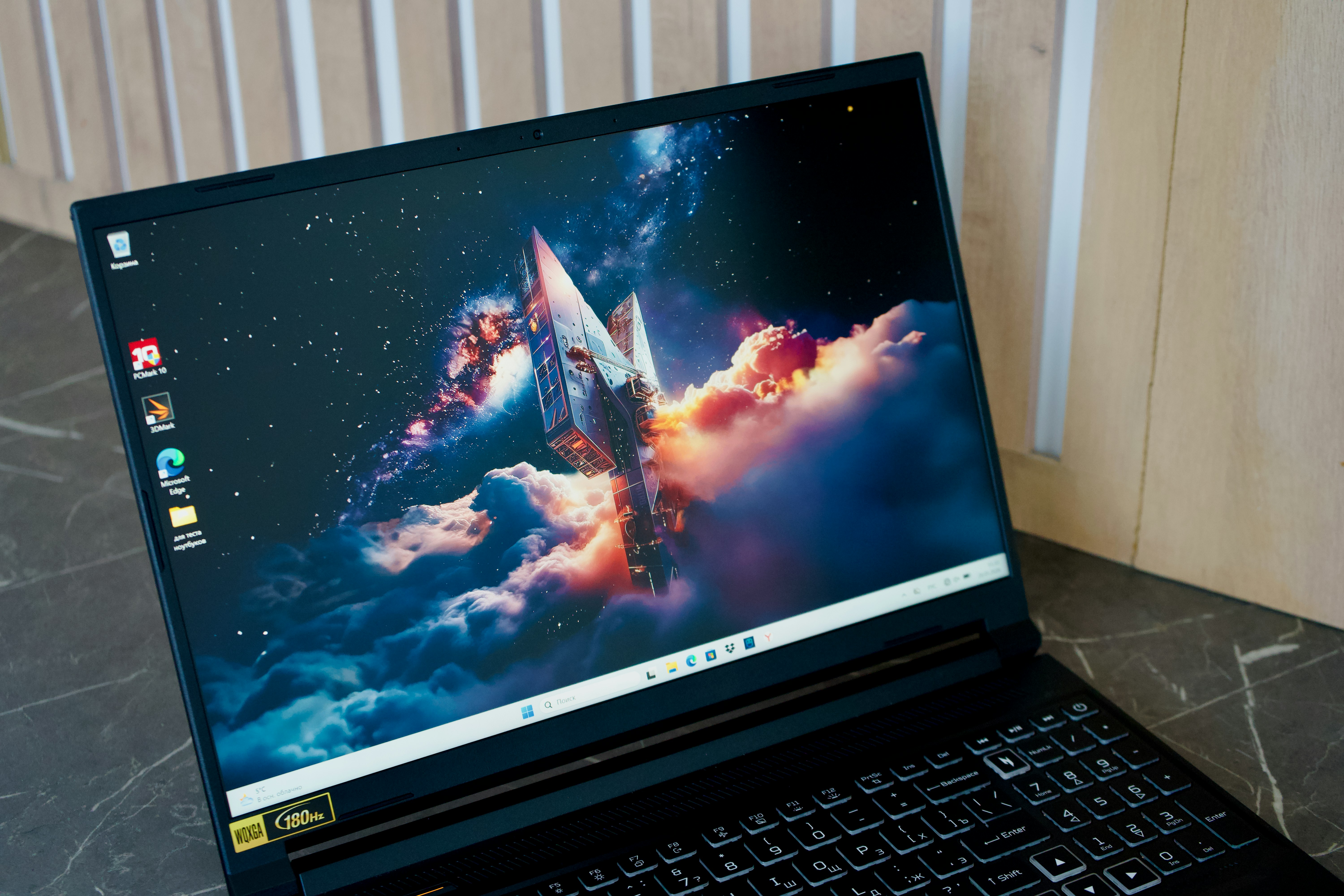 Low-angle shot of a modern laptop displaying a vibrant space desktop with nebulae and rocket imagery, resting on a dark surface in a softly lit room. The scene highlights the screen’s color depth and sleek design.