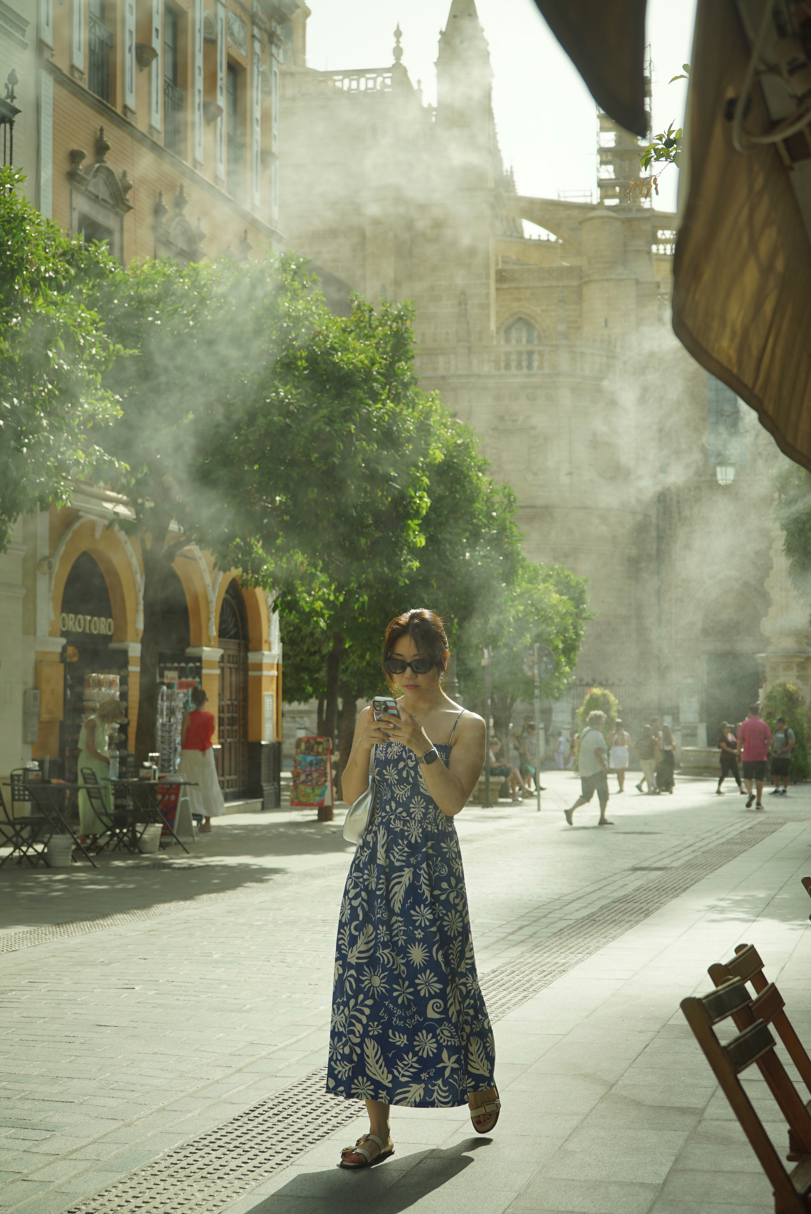 Cooling water mist drifts through a lively pedestrian street near Seville Cathedral as a visitor checks a smartphone during a hot Andalusian summer afternoon. Shot with Sony a7s // Una fina niebla refrescante recorre una calle peatonal cercana a la Catedral de Sevilla mientras una visitante consulta su teléfono en una calurosa tarde de verano.