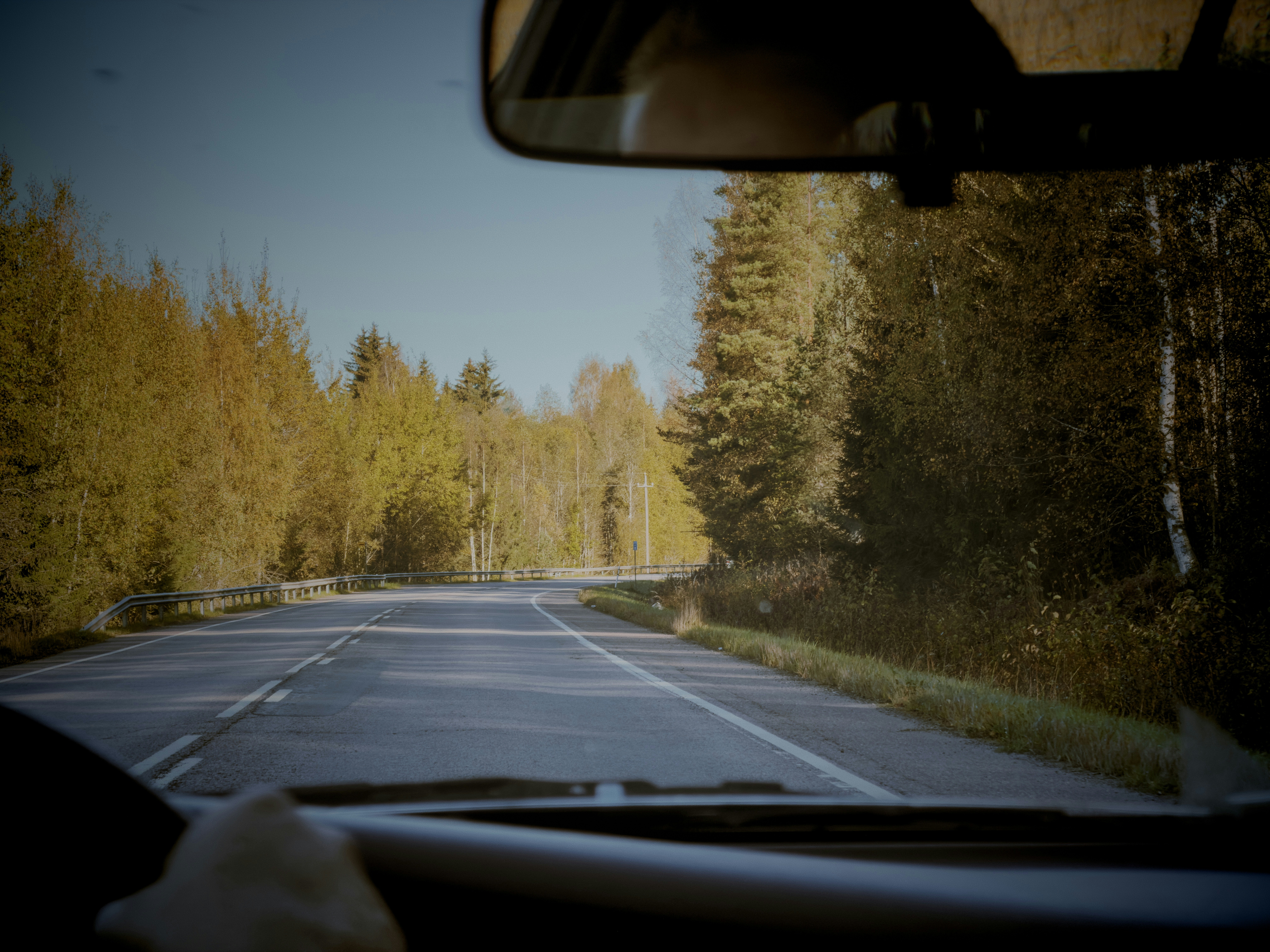 Autumn trees line a winding road viewed from inside a car, with dashboard edges framing the scene and a clear sky above.